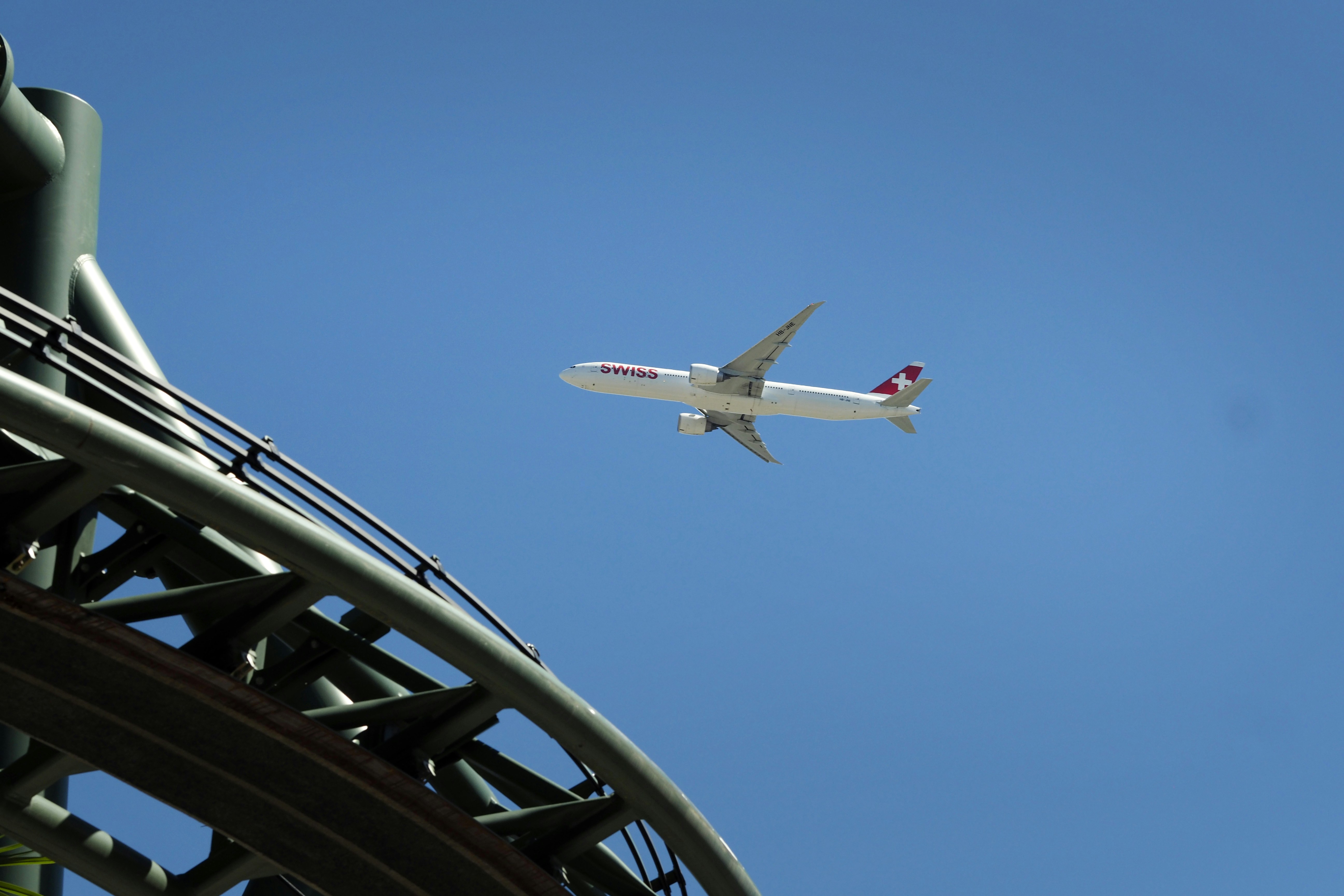 An airplane flies overhead against a bright blue sky with a partial foreground structure framing the lower left. The image emphasizes scale and motion with a clean, minimal backdrop.