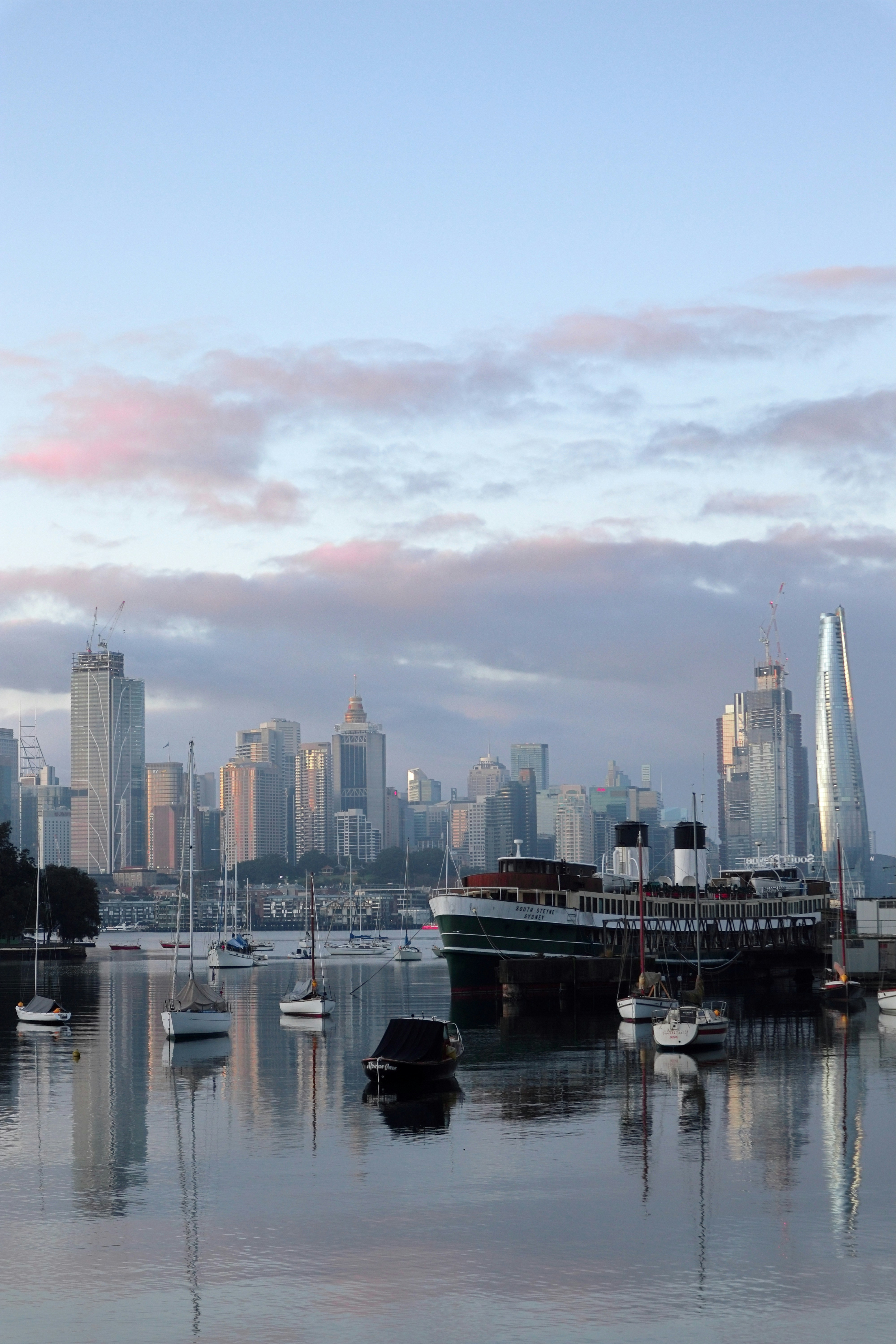 Historic ship docked alongside modern skyscrapers, with sailboats gently bobbing in the calm water at dawn.