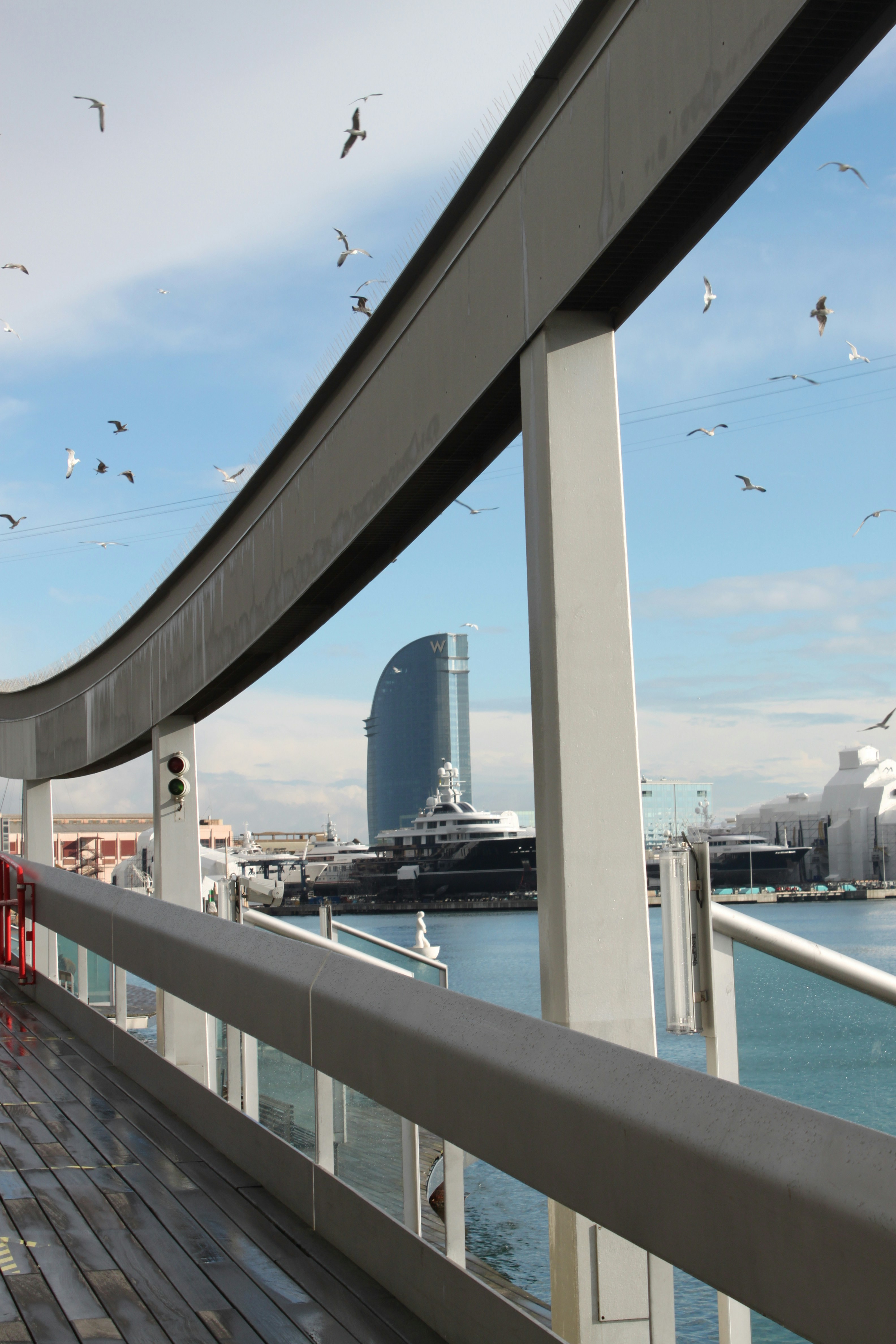 A modern architectural walkway curves gracefully over a serene harbor, with a luxury yacht and city skyline in the background. Seagulls soar above, adding life to the scene.