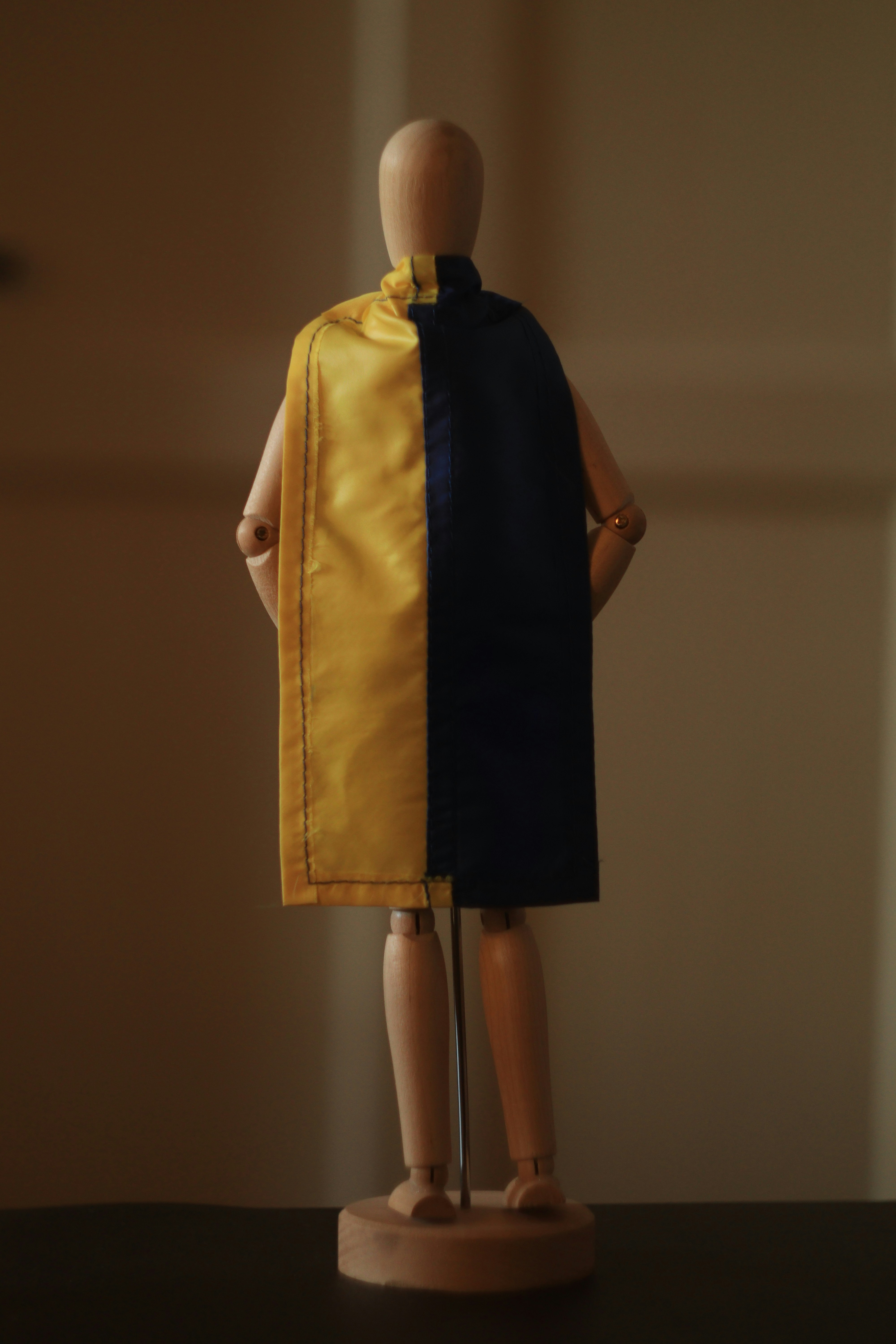 Wooden mannequin adorned with a yellow and blue cape, standing against a softly lit background.