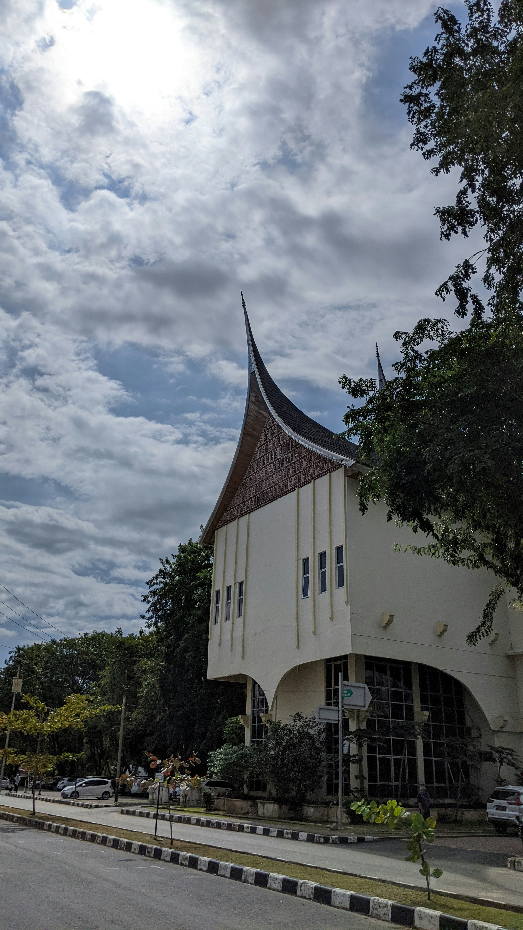 Architectural building with a dramatic sail-shaped roof rises beside a tree-lined street under a bright, cloud-filled sky.