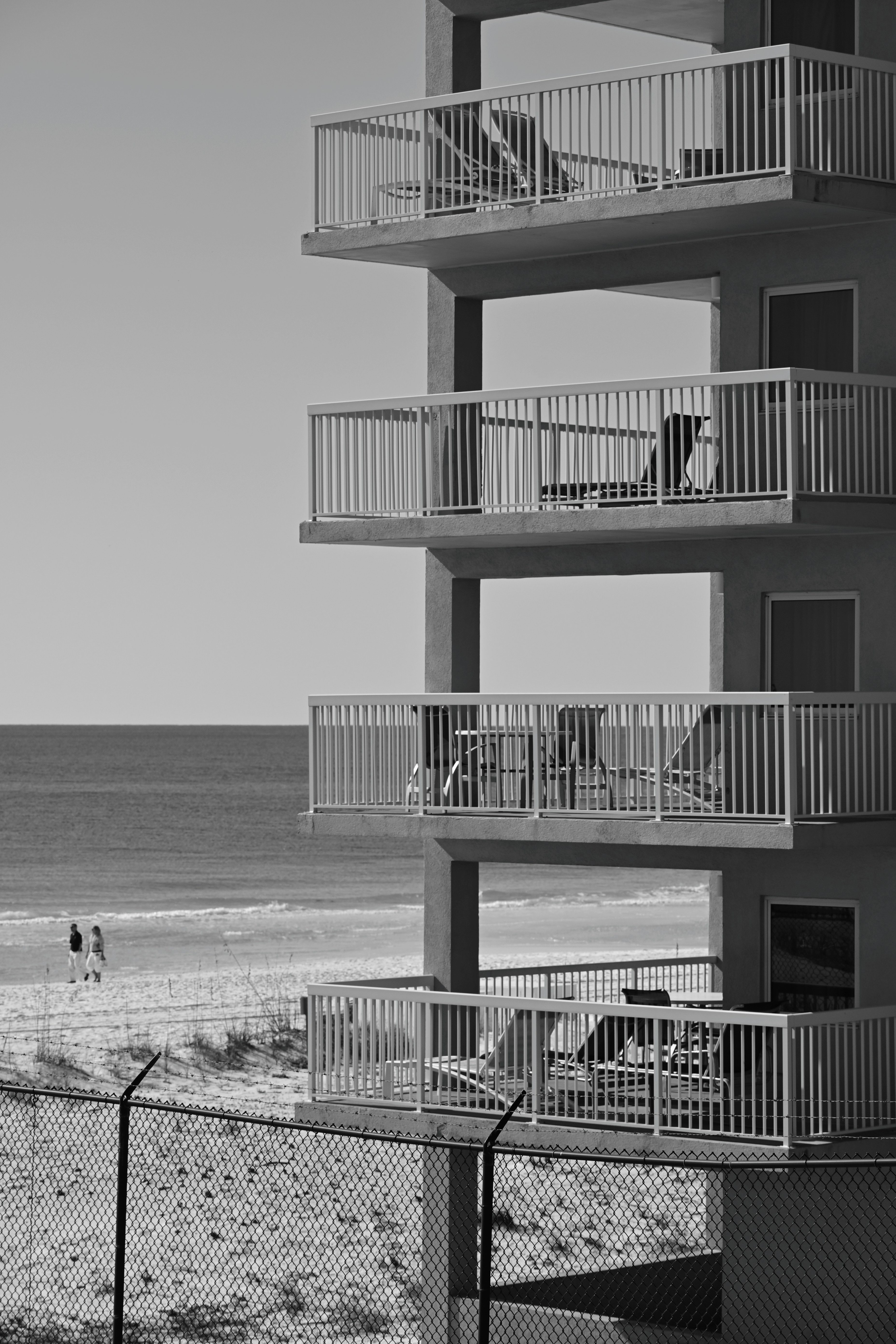 Modern beachfront condominium with balconies overlooking the ocean, framed by a sandy shore and distant beachgoers. 