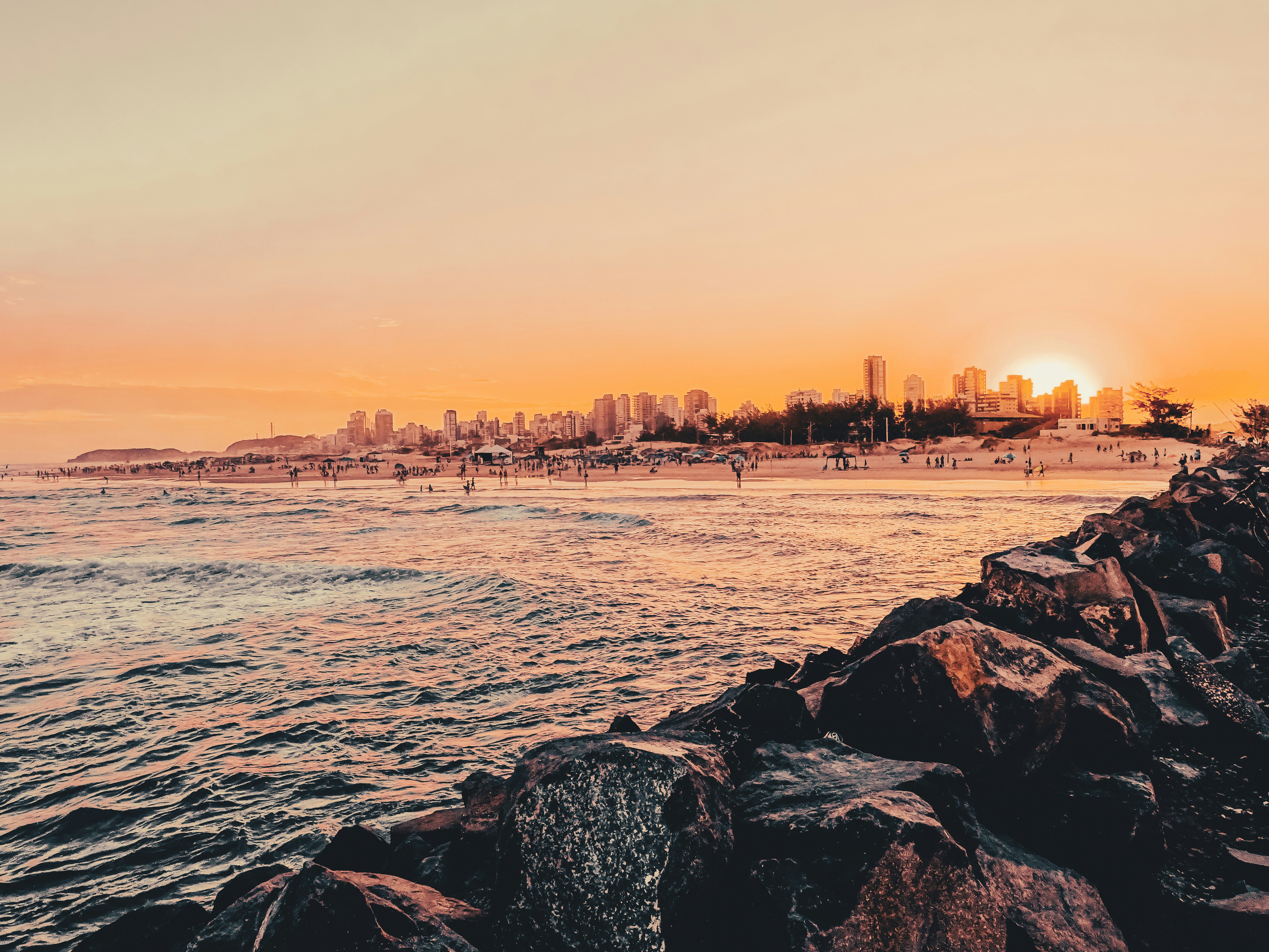 Golden sunset casting warm hues over a beachside cityscape, with gentle waves lapping against rocky shores.