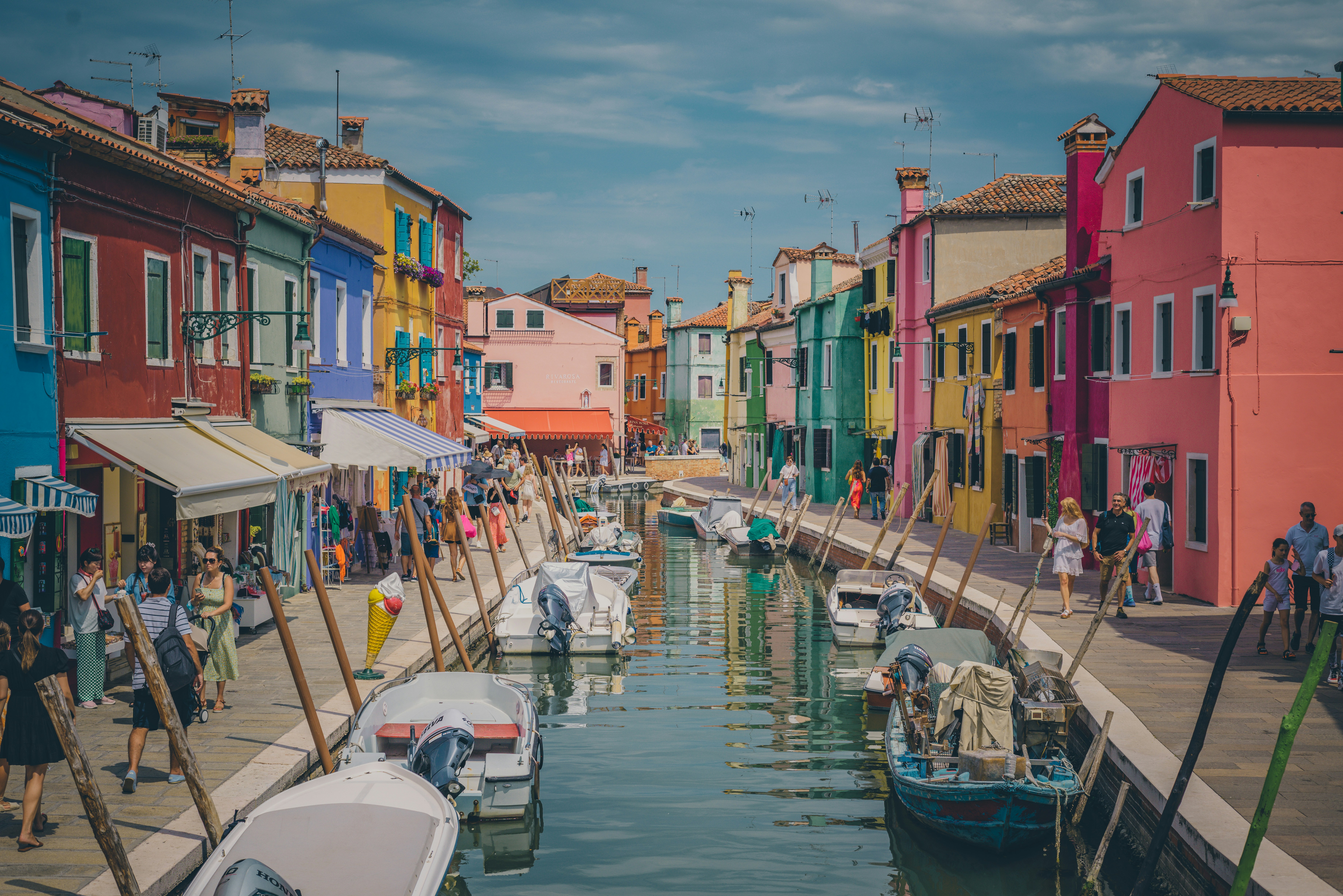 Colorful buildings line the canal of Burano, with boats moored along the banks and visitors strolling the vibrant streets.