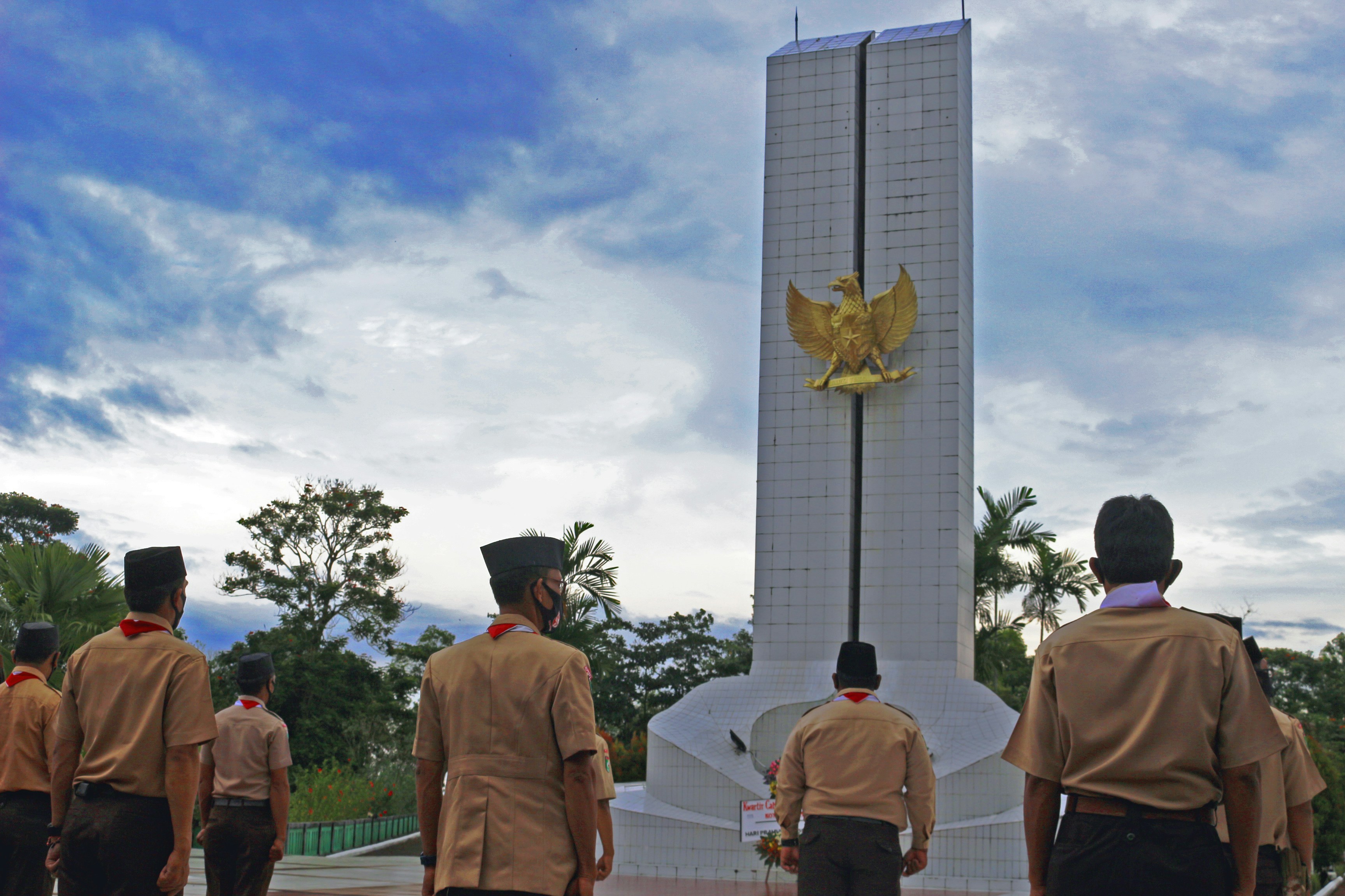 Uniformed individuals stand before a tall monument with a golden emblem against a partly cloudy sky.