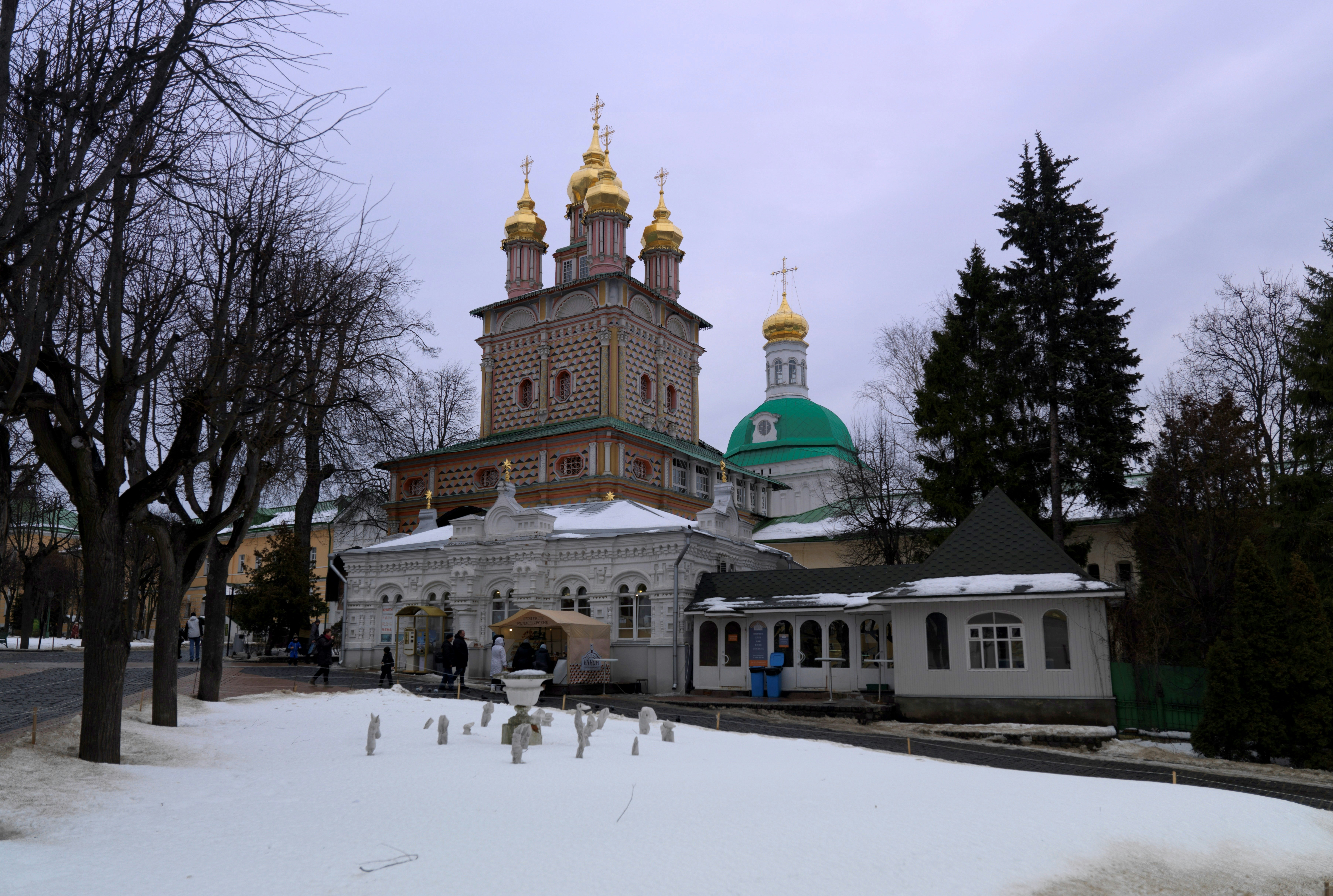 Ornate building with golden domes and a tower, surrounded by snow and bare trees in a serene winter setting.