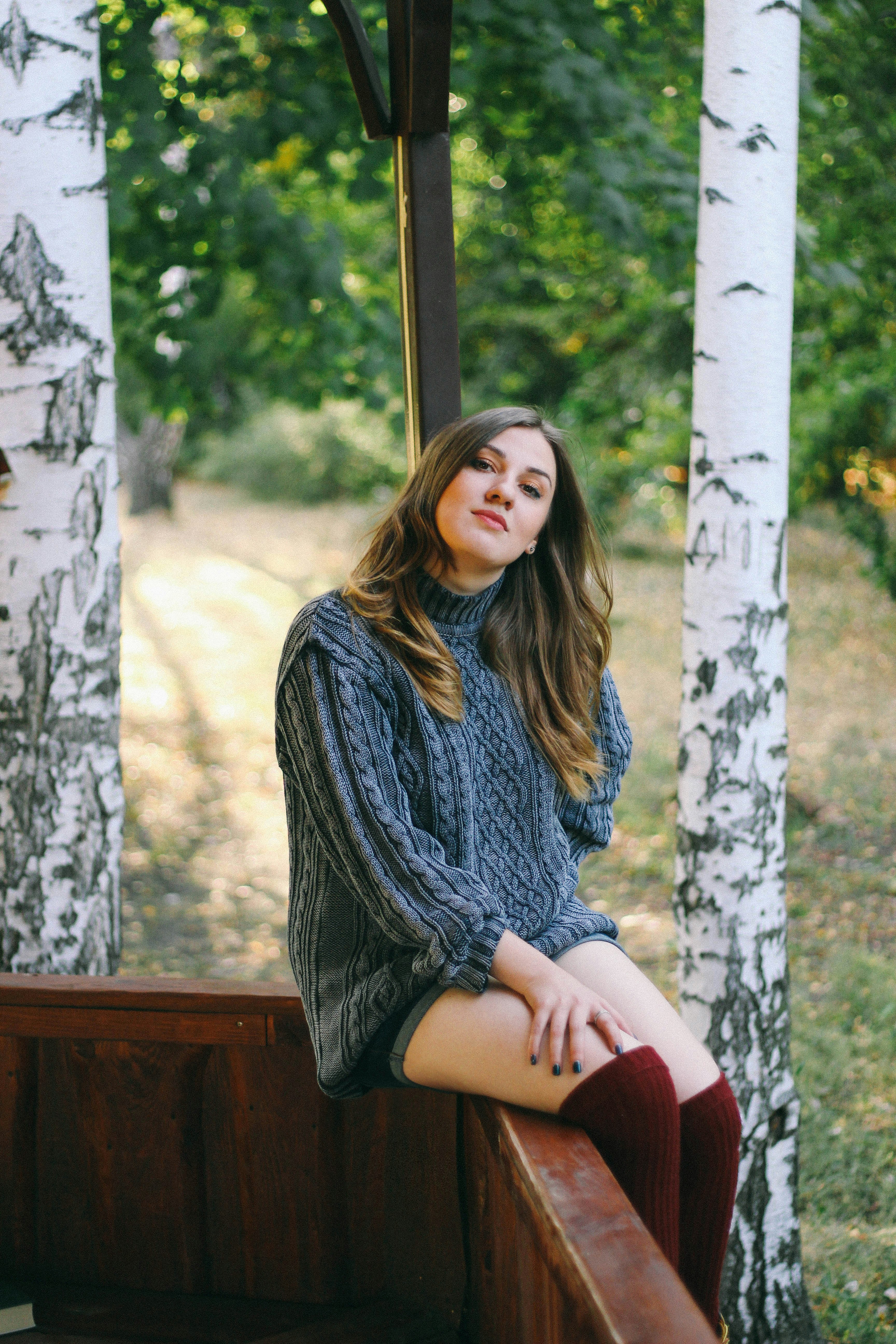 Young woman in a cozy sweater and knee-high socks sitting on a wooden ledge surrounded by birch trees, with soft sunlight filtering through the leaves.