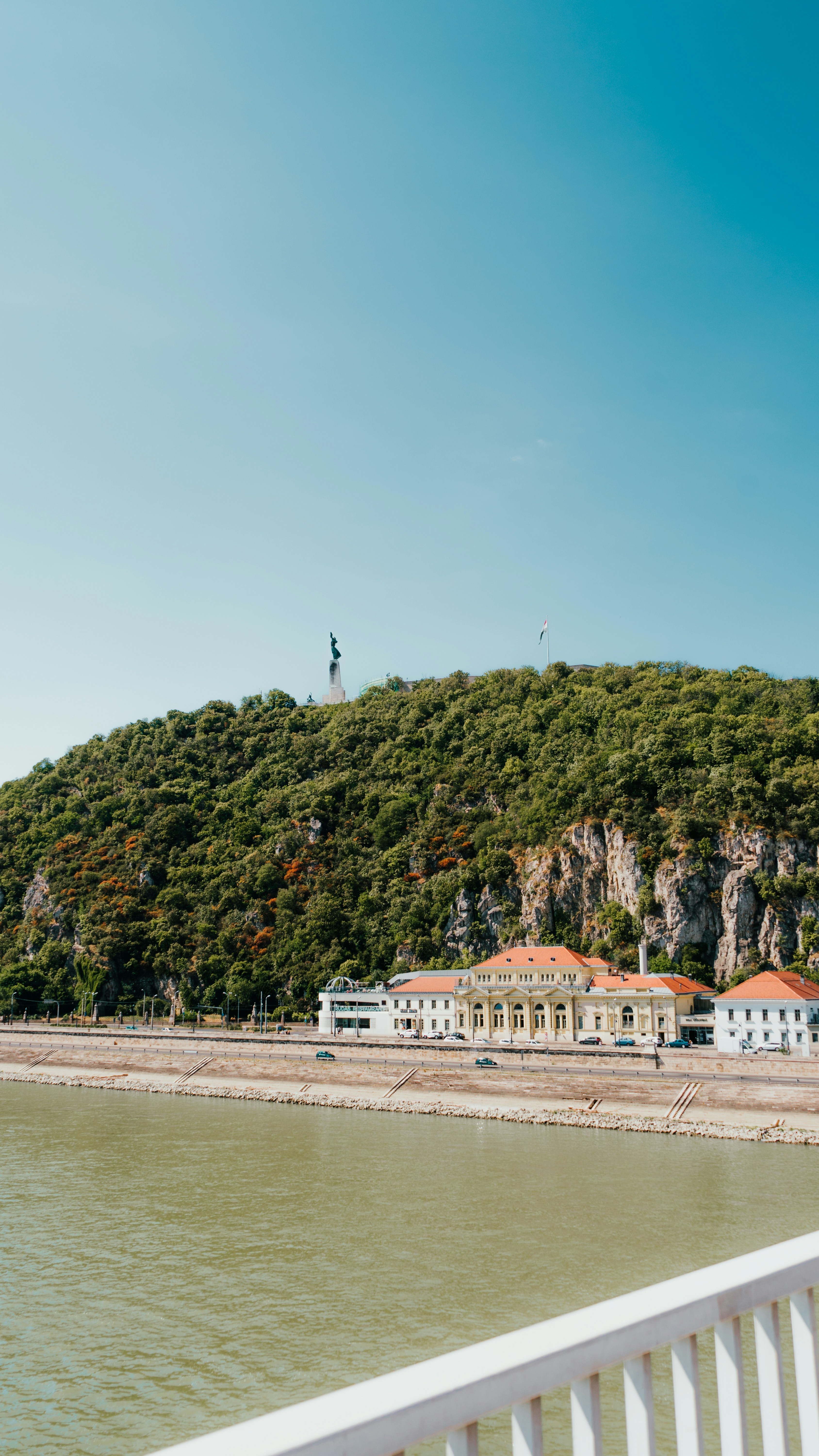 Photograph of a riverfront with a forested hill, a stately building along the bank, and a statue atop the hill under a clear blue sky.