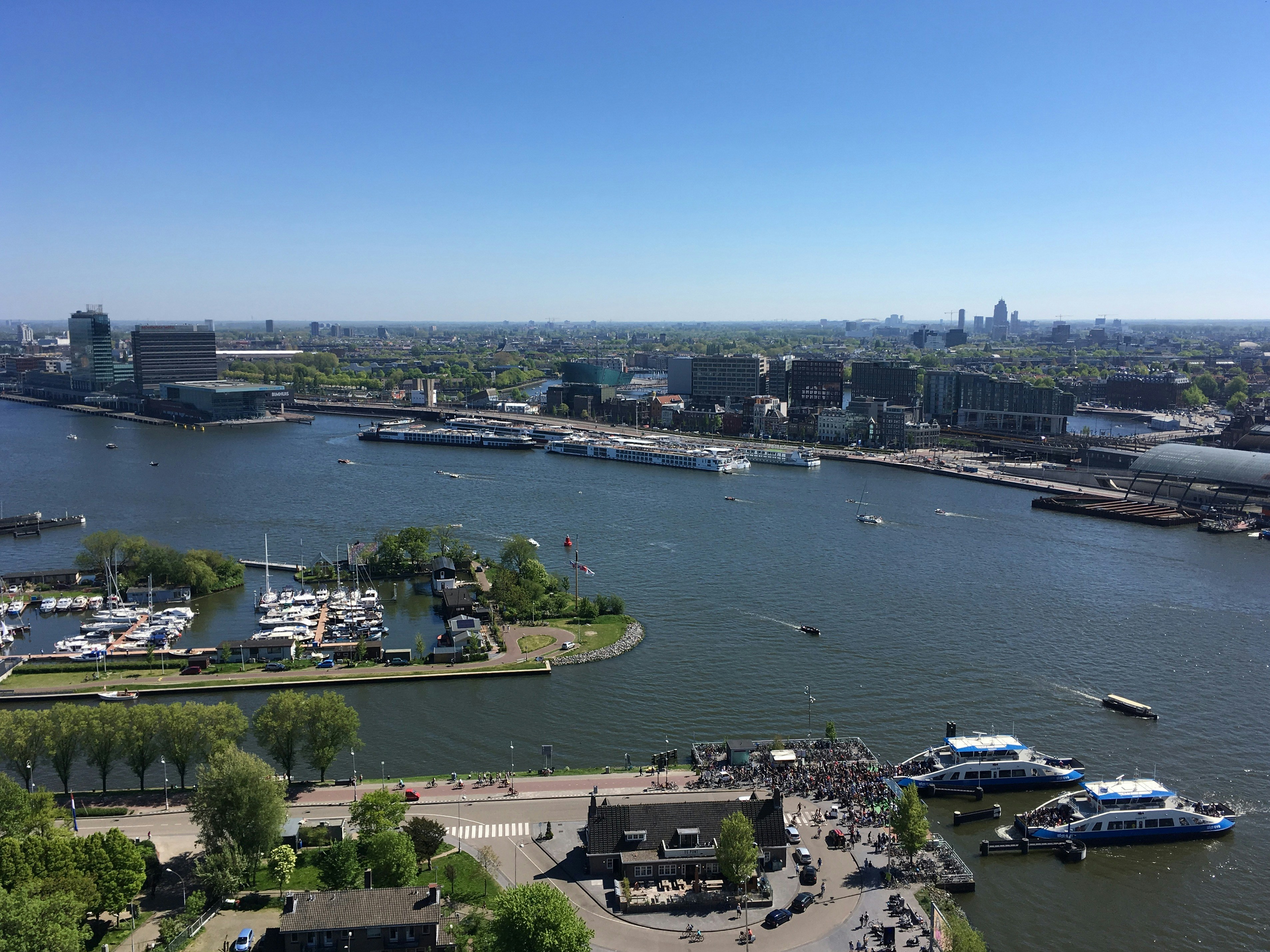 Aerial view of a bustling harbor with boats and green parks lining the riverbank. The city skyline stretches in the background under a clear blue sky.