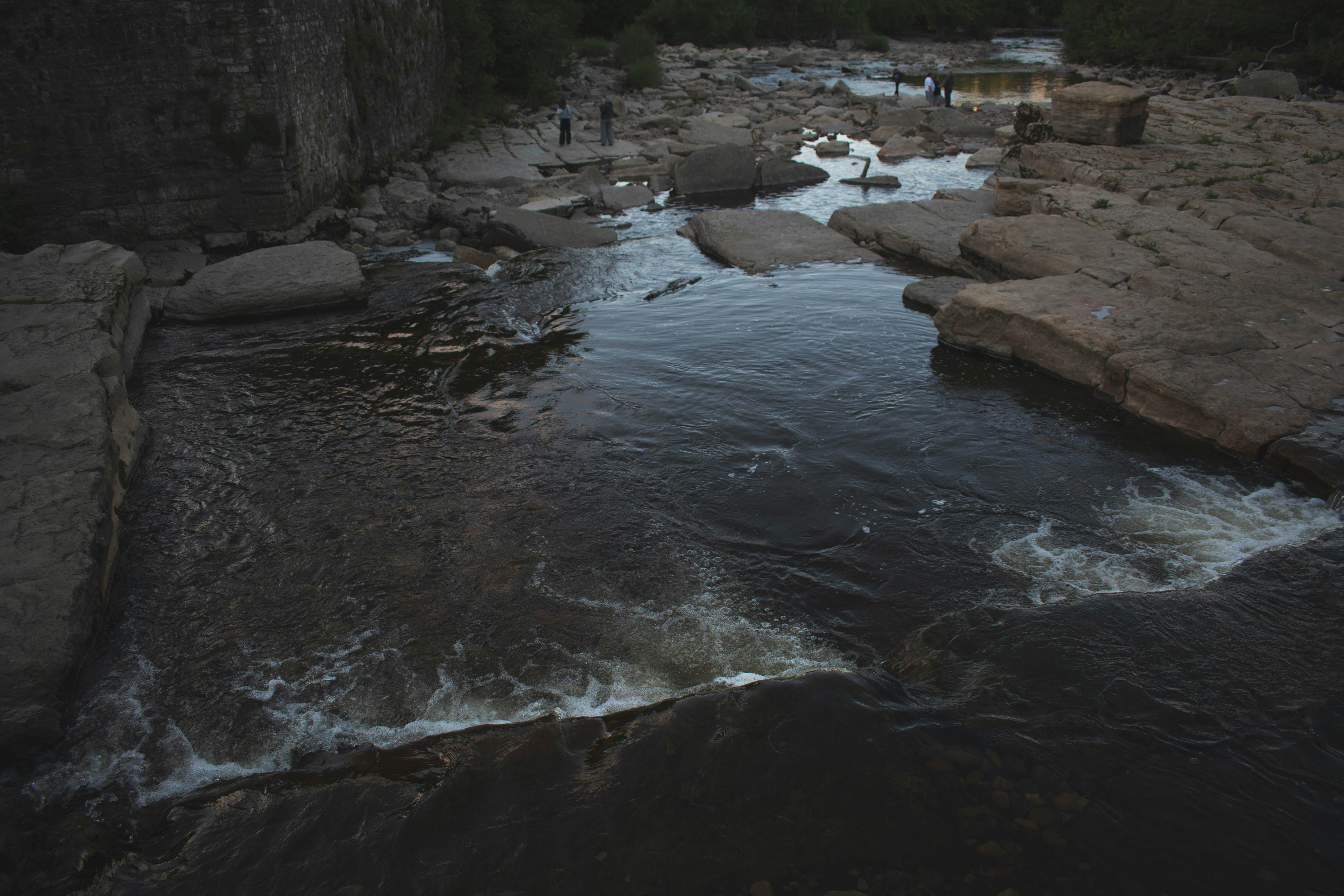 Gentle river flowing over rocky terrain with silhouettes of people in the background, depicting a serene natural setting.