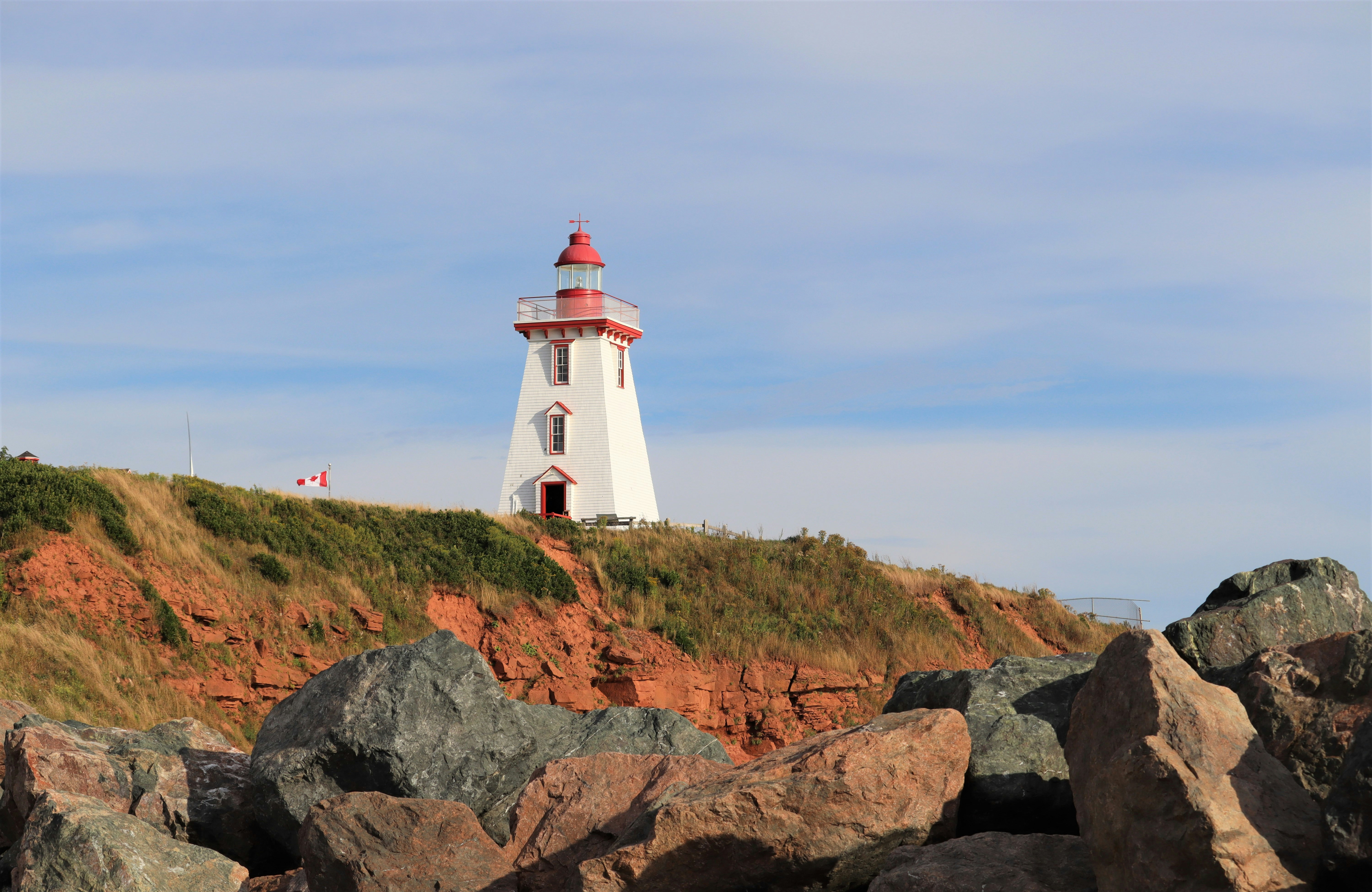 A classic lighthouse stands proudly atop a rocky cliff, framed by lush greenery and a clear blue sky. The Canadian flag waves gently nearby.