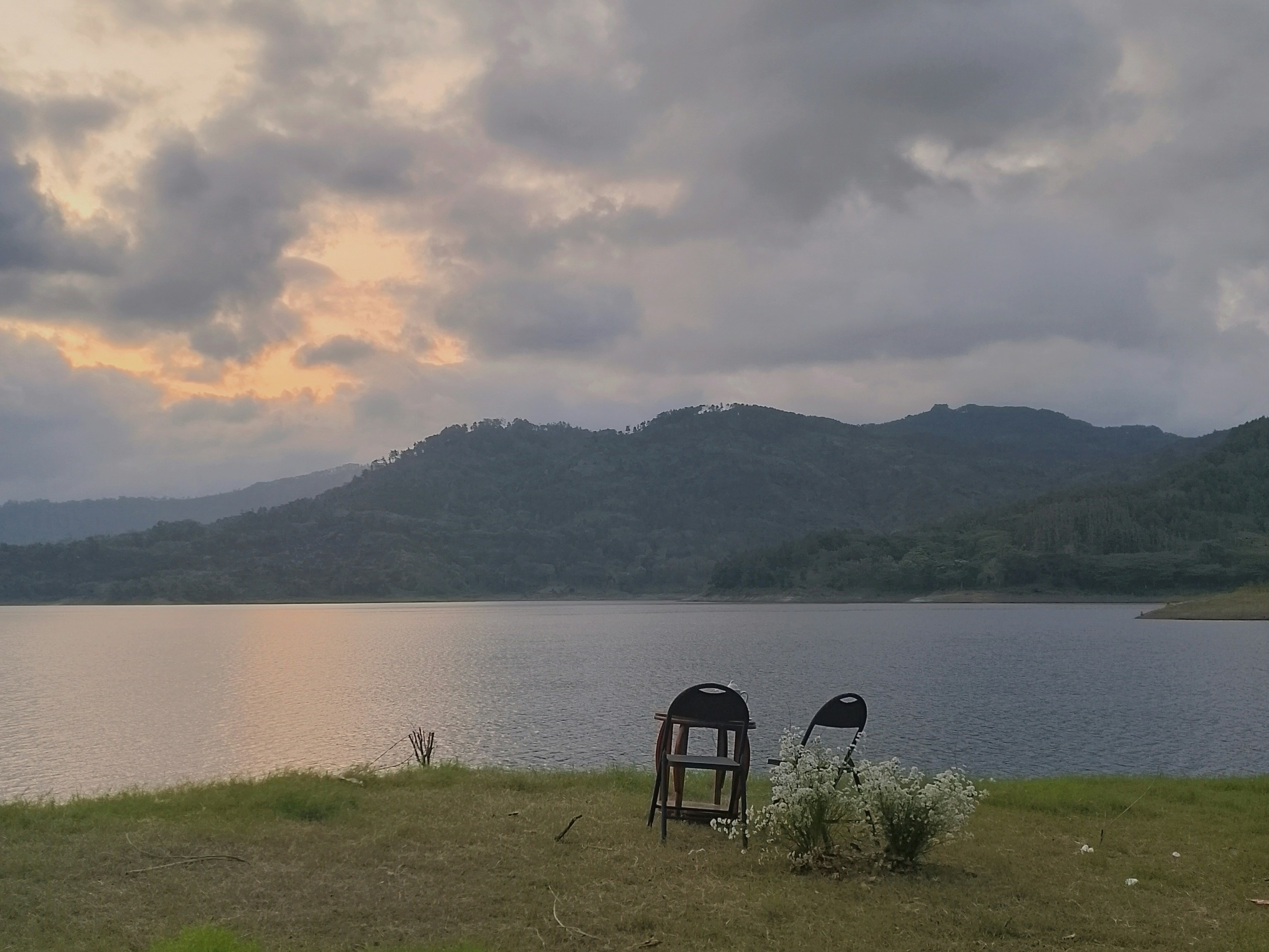 Two empty chairs beside a tranquil lake at dusk, framed by distant mountains and a cloudy sky. 