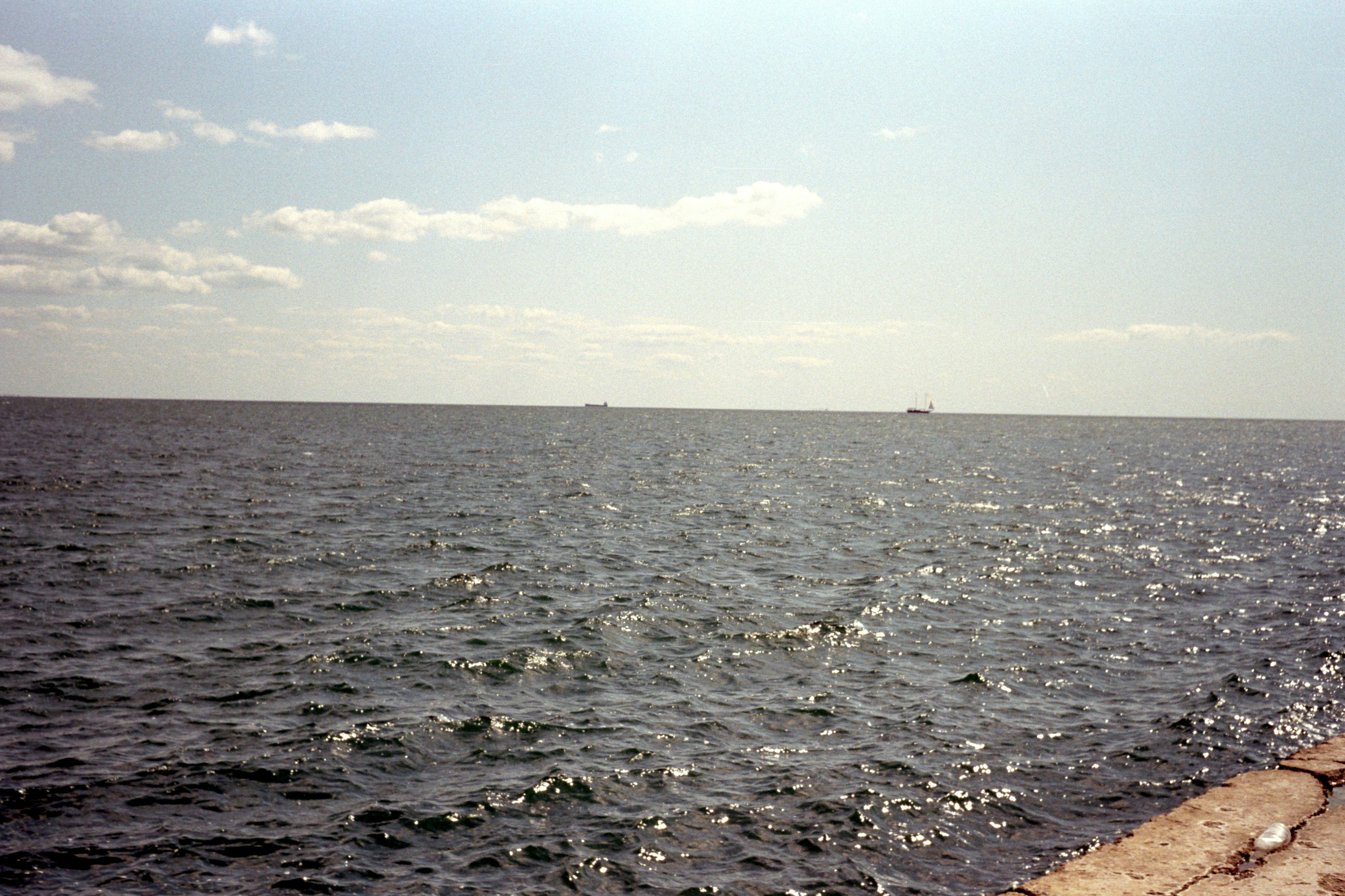 Expansive view of a calm sea under a bright sky, with distant ships barely visible on the horizon. The shimmering water reflects the sunlight beautifully.