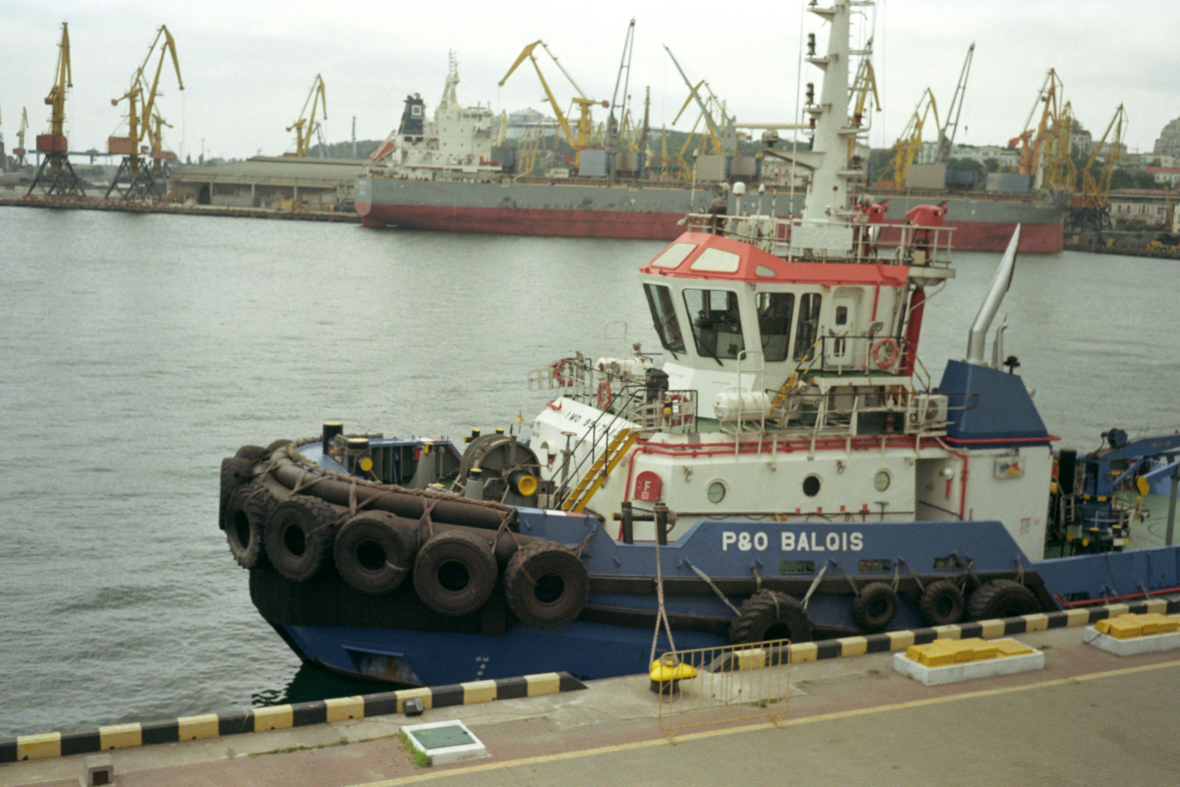 Tugboat P80 BALOIS maneuvering in a busy harbor with cargo ships in the background. The scene captures the essence of maritime operations.