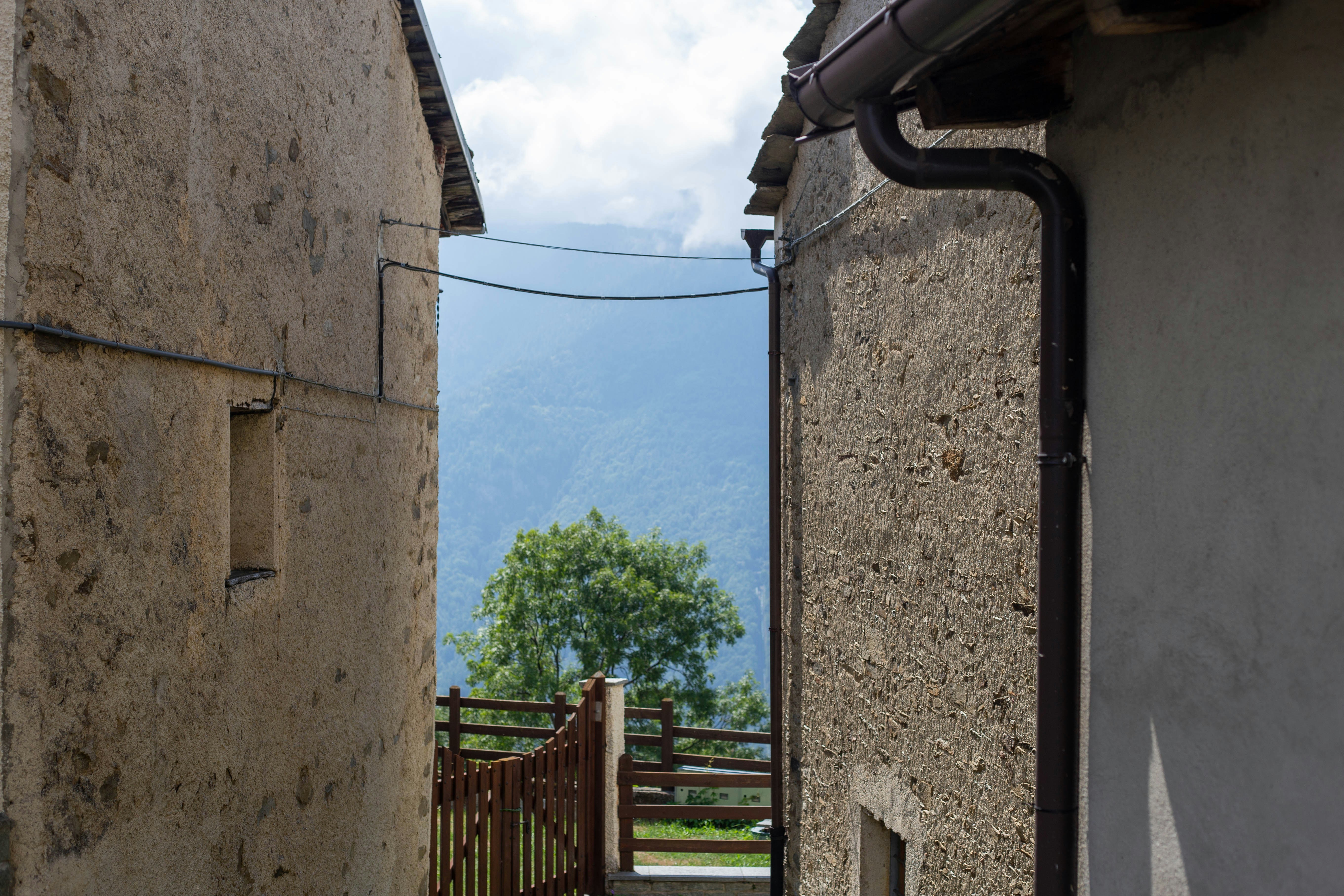 Narrow alley between two stone buildings leading to a distant view of a tree against a mountainous backdrop under a partly cloudy sky.