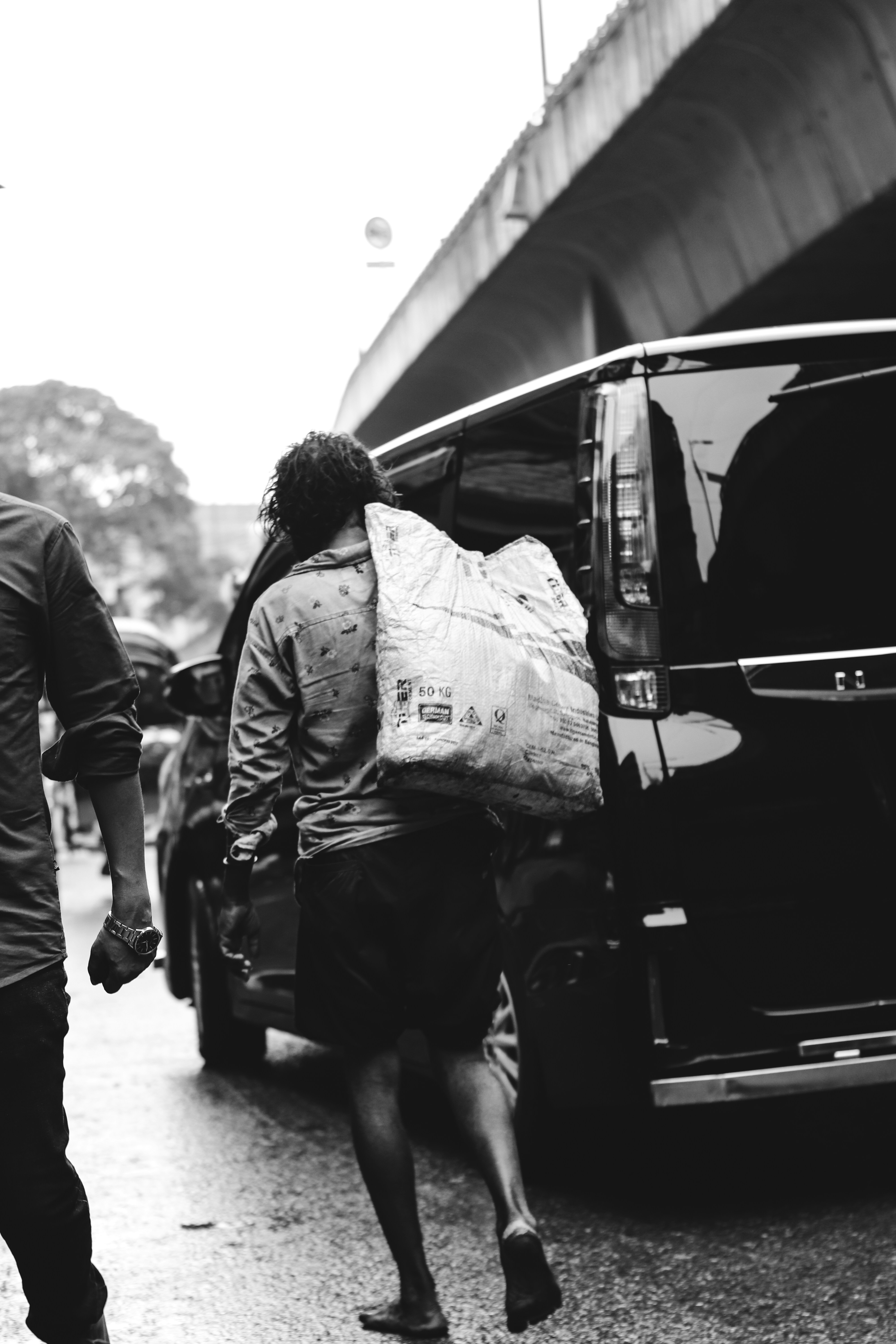 Monochrome Street Run: A Bag-Laden Pedestrian Moving Past a Van Under the Bridge