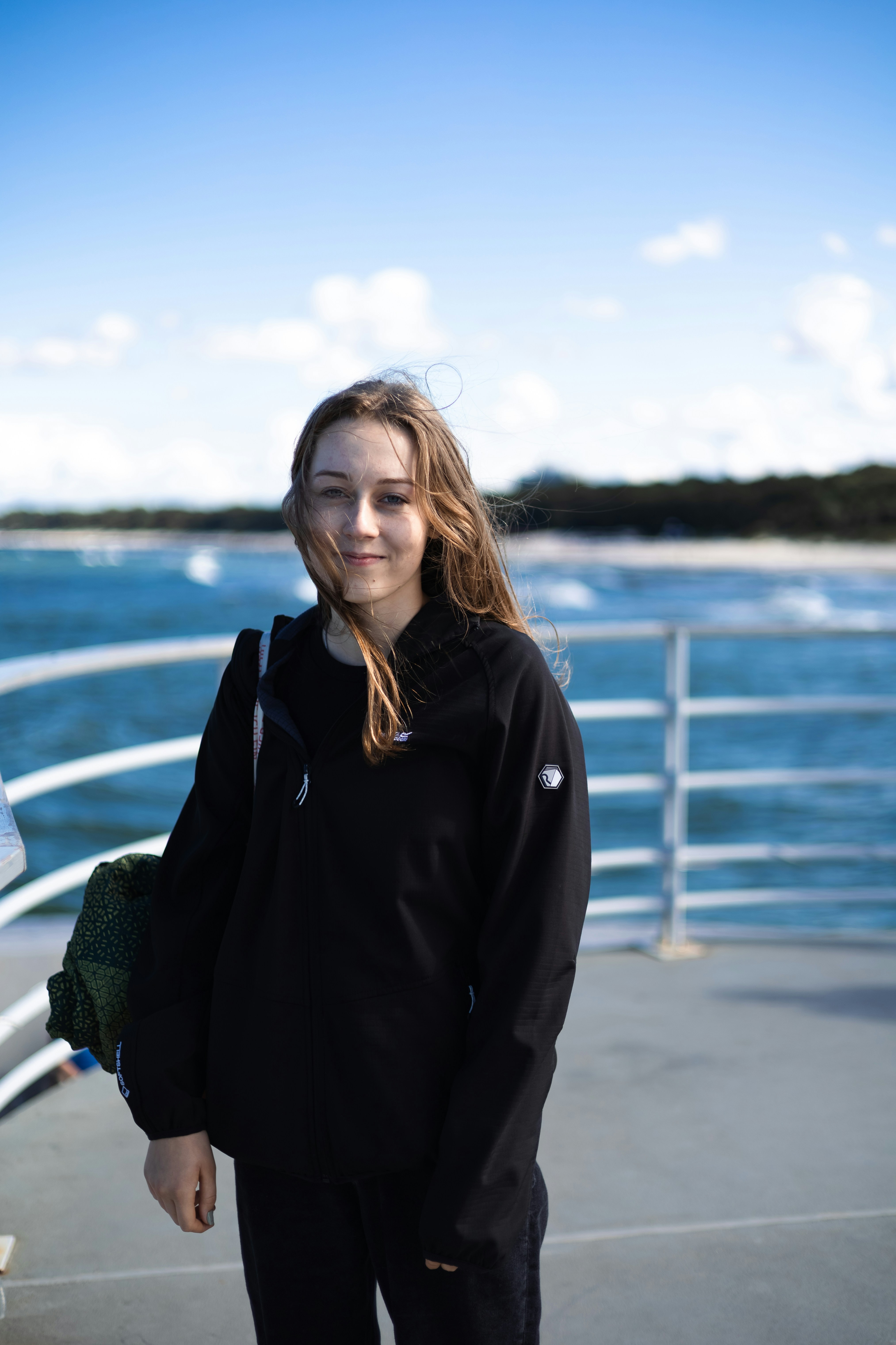 Young woman standing on a pier with wind-swept hair, overlooking the ocean waves. The scene captures the essence of a coastal outing.