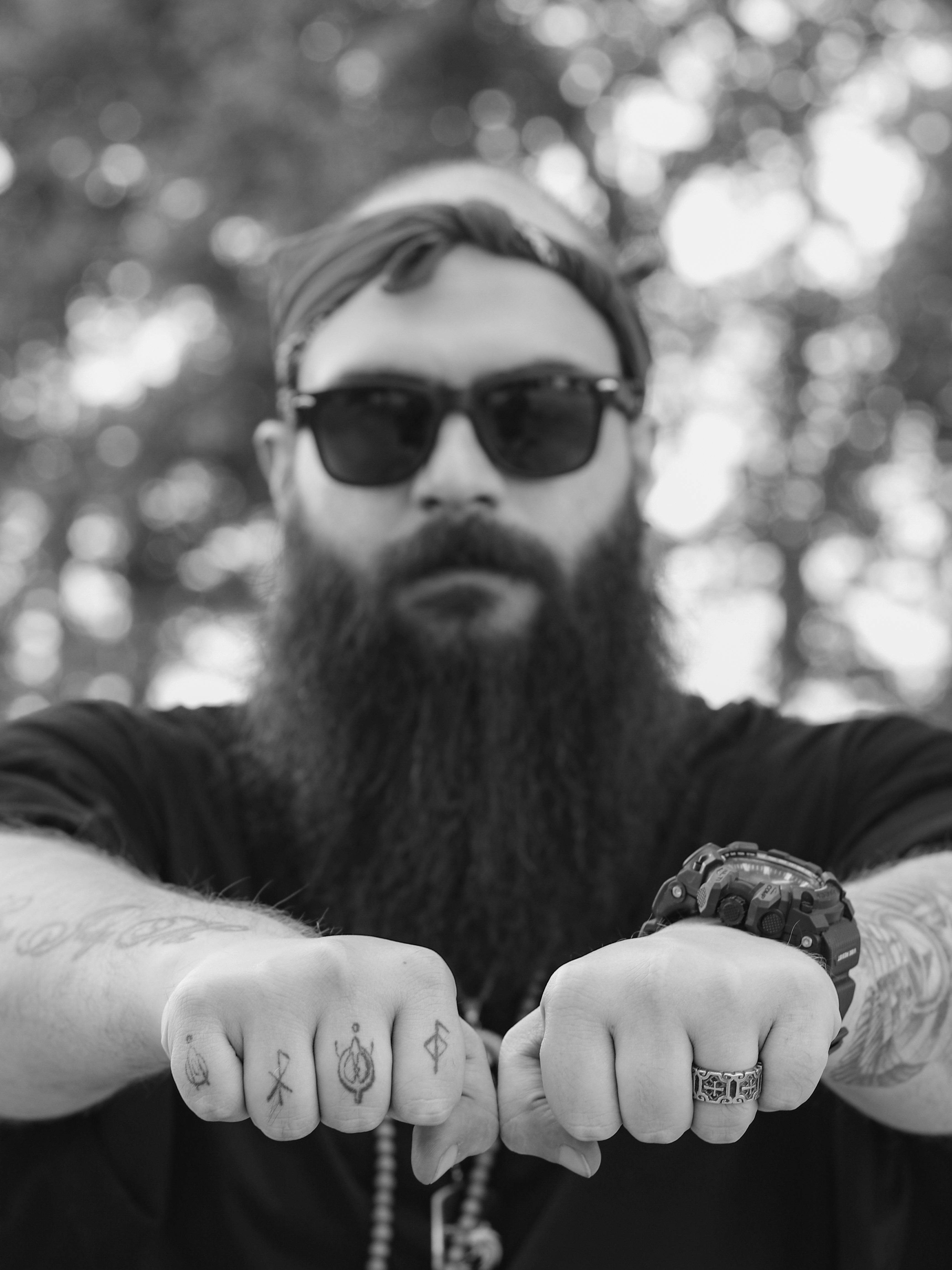 A bearded man showcases intricate tattoos on his hands while wearing sunglasses, set against a softly blurred background. The monochrome tones enhance the focus on his detailed ink.