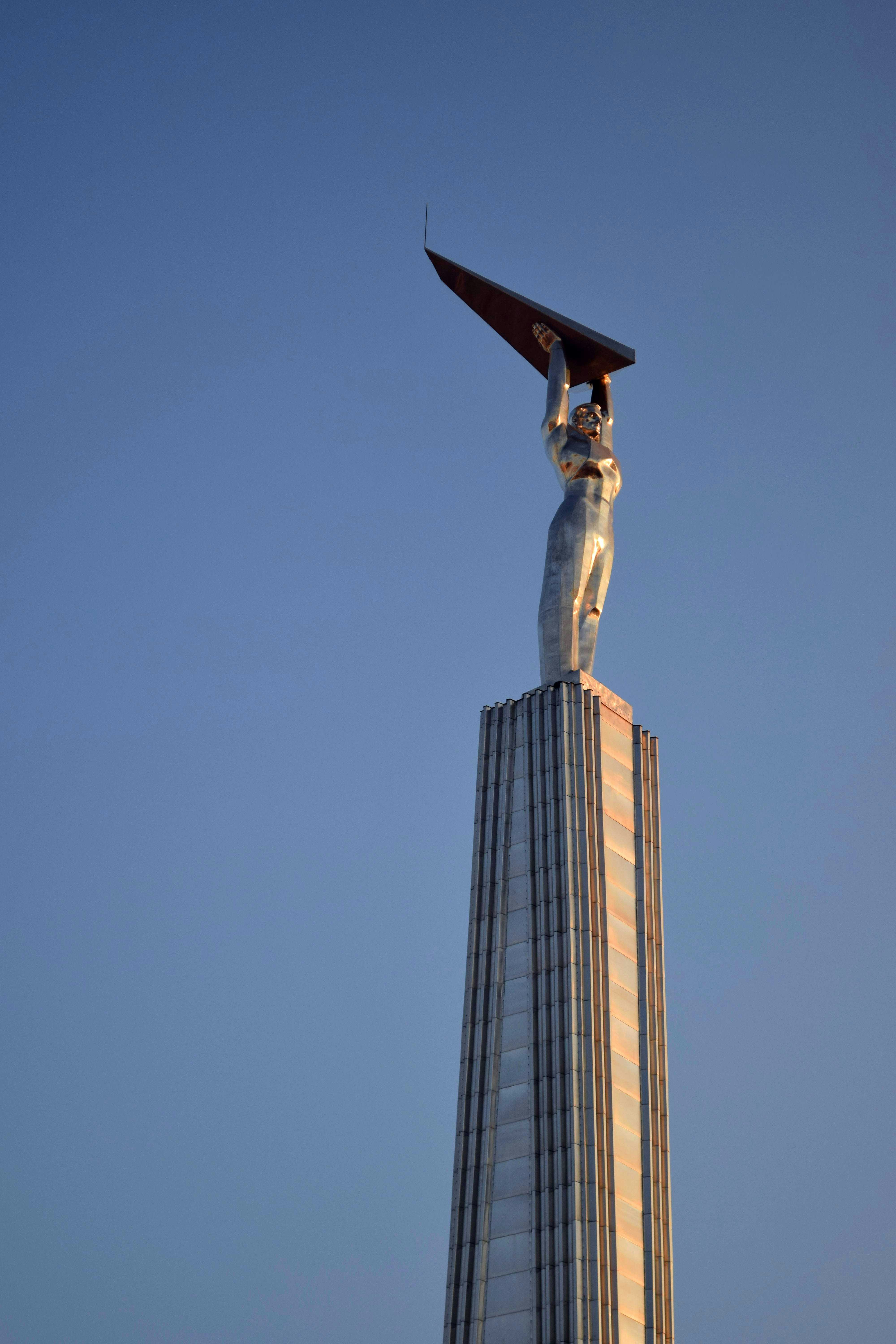 A towering silver statue of a figure holding a model airplane aloft, symbolizing progress in aviation against a clear blue sky.