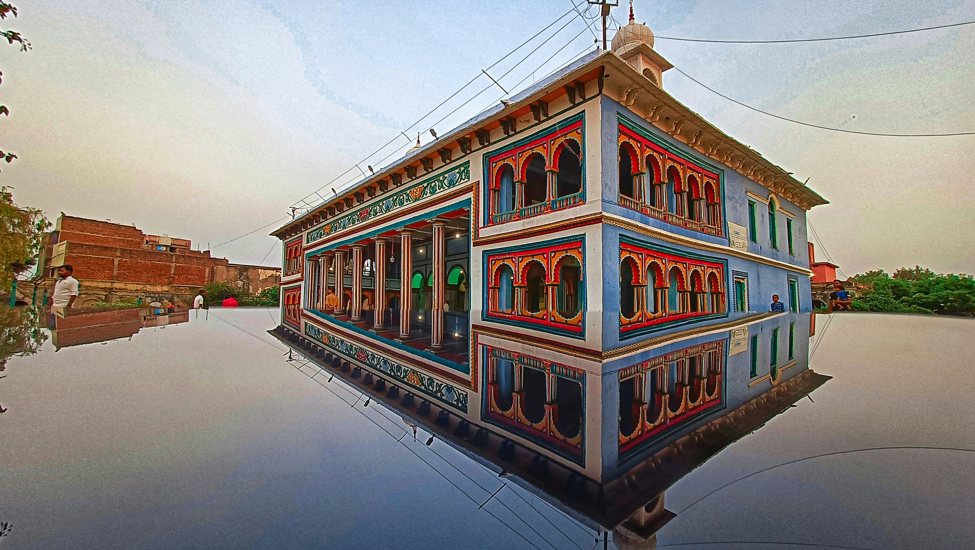 Temple with colorful arches reflected perfectly in a smooth water surface, surrounded by greenery and clear sky.
