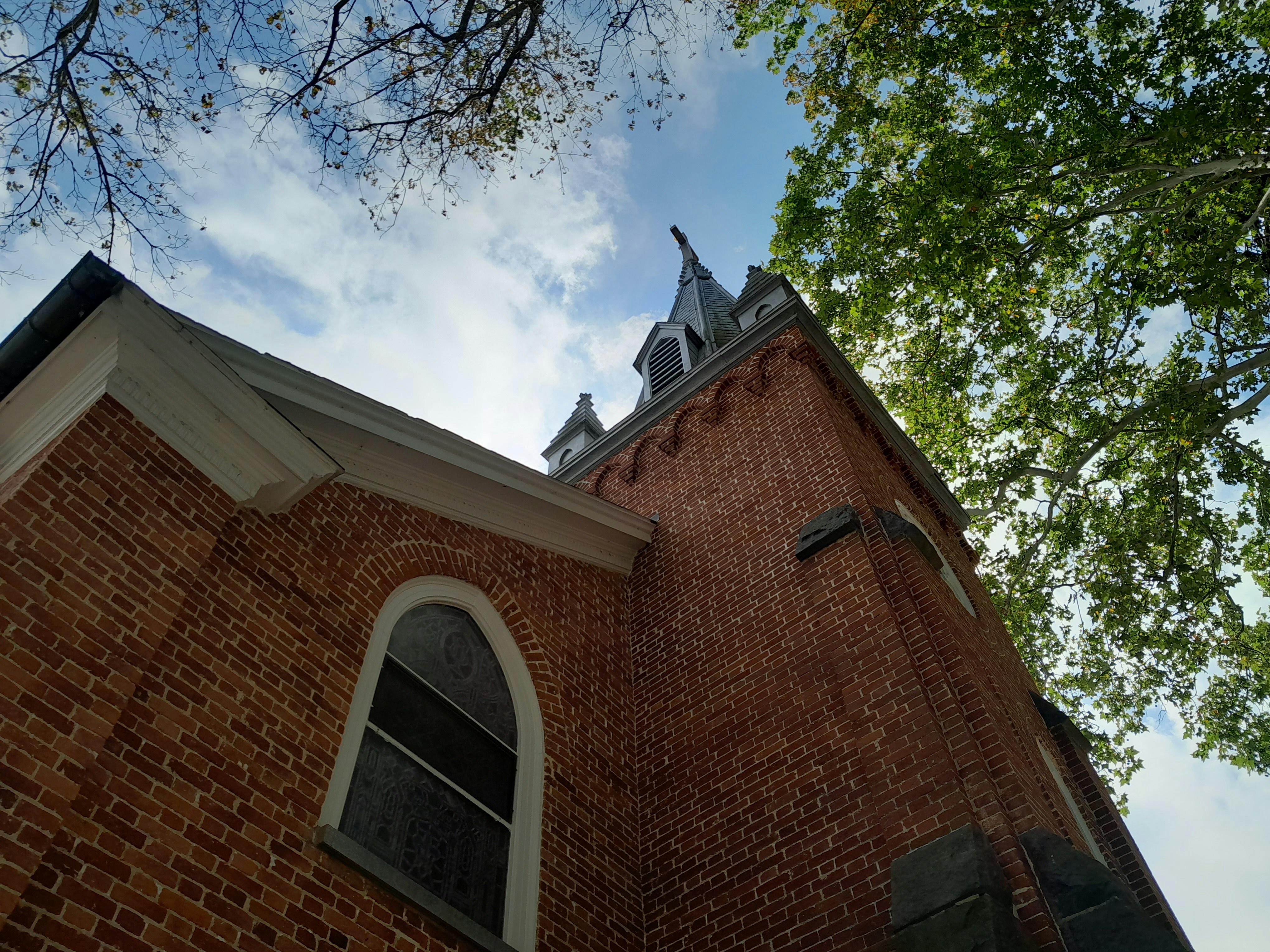 Historic brick church viewed from below, showcasing its architectural details against a backdrop of leafy trees and a cloudy sky.