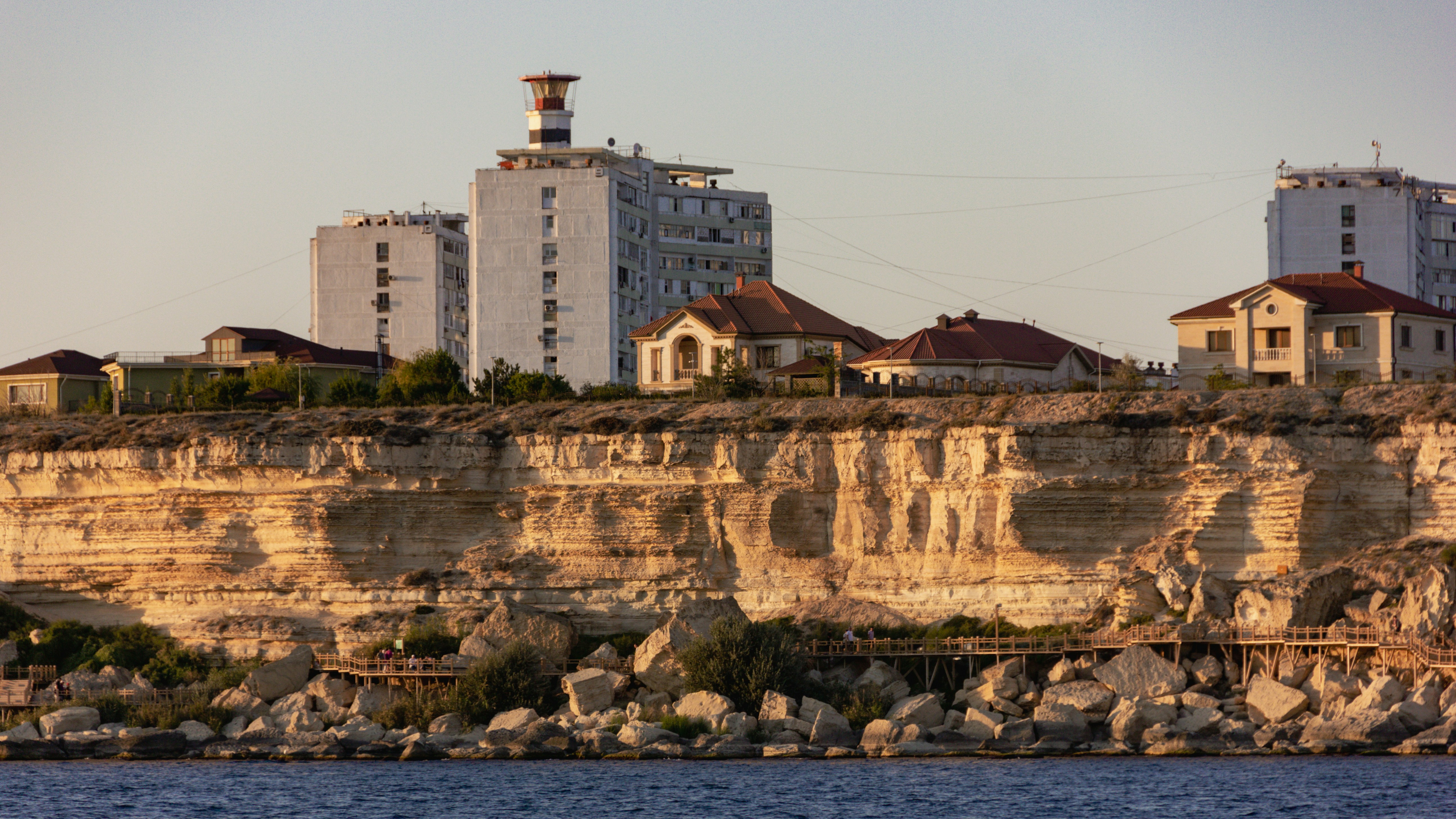 Buildings perched atop rugged cliffs overlooking a serene body of water under warm sunset light.