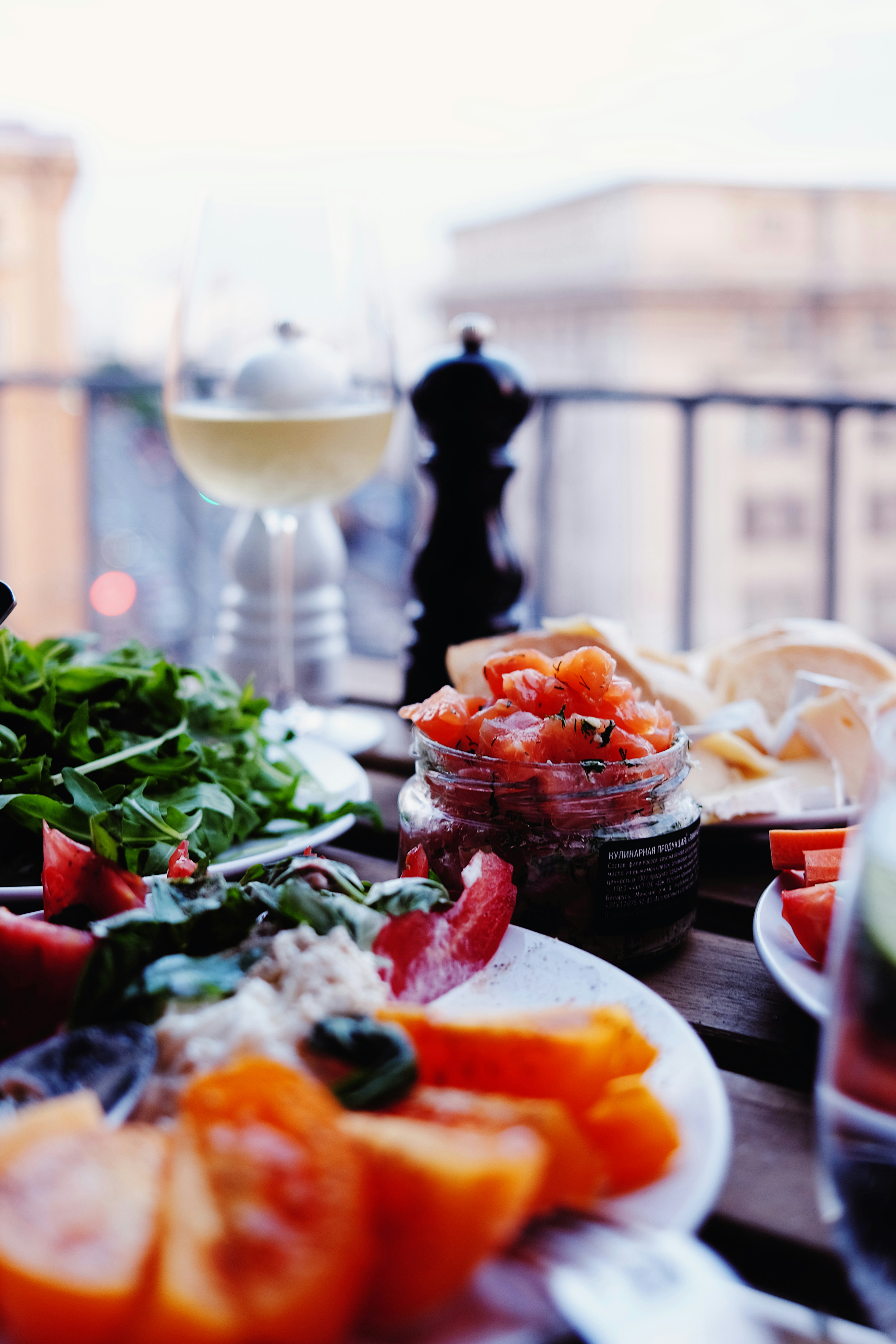 An elegant spread of fresh ingredients and gourmet delights on a balcony, with a glass of white wine in the background. The soft evening light enhances the vibrant colors of the meal.