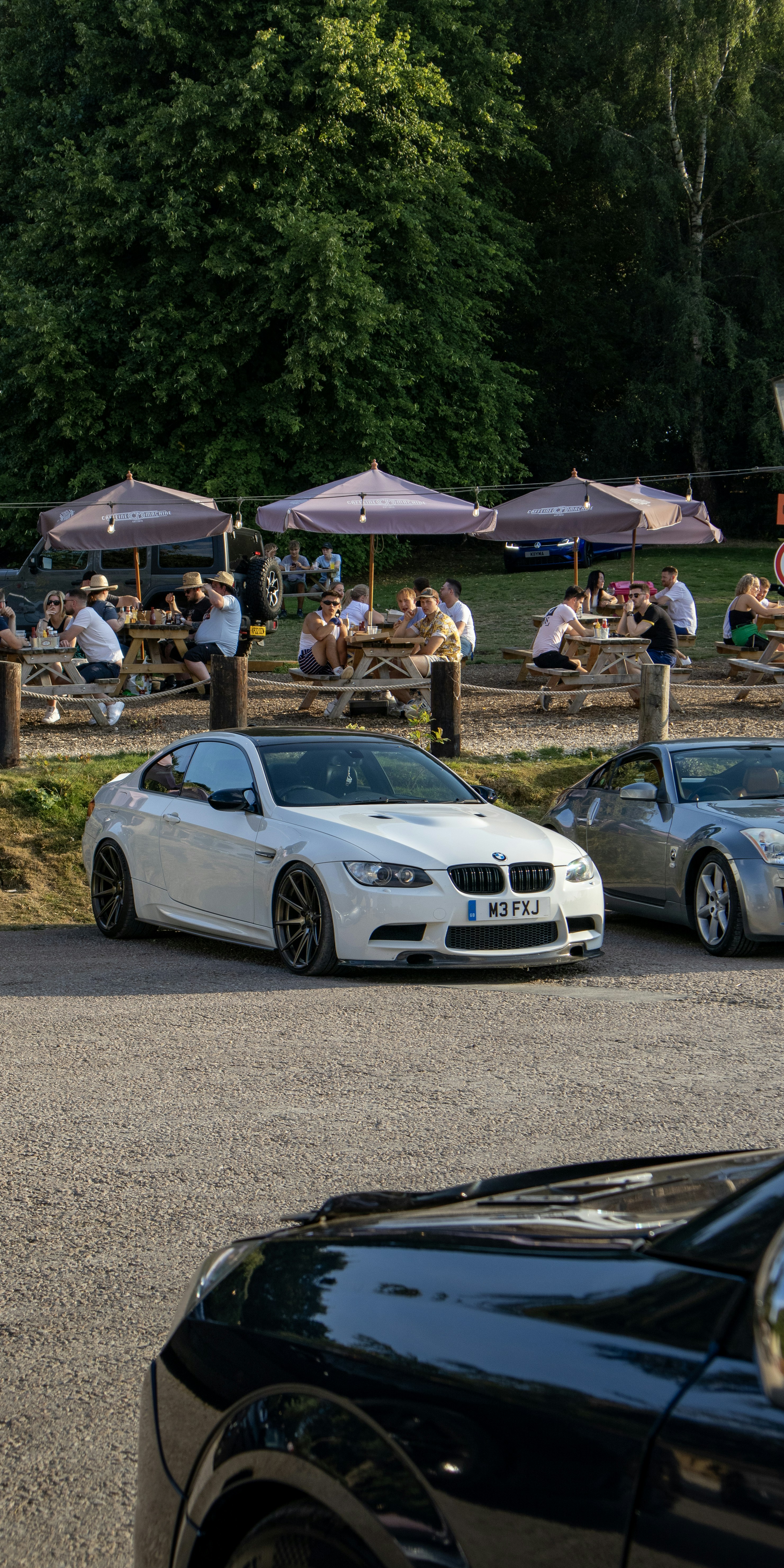 Sunlit White BMW Framed by a Bustling Outdoor Café Under Purple Umbrellas