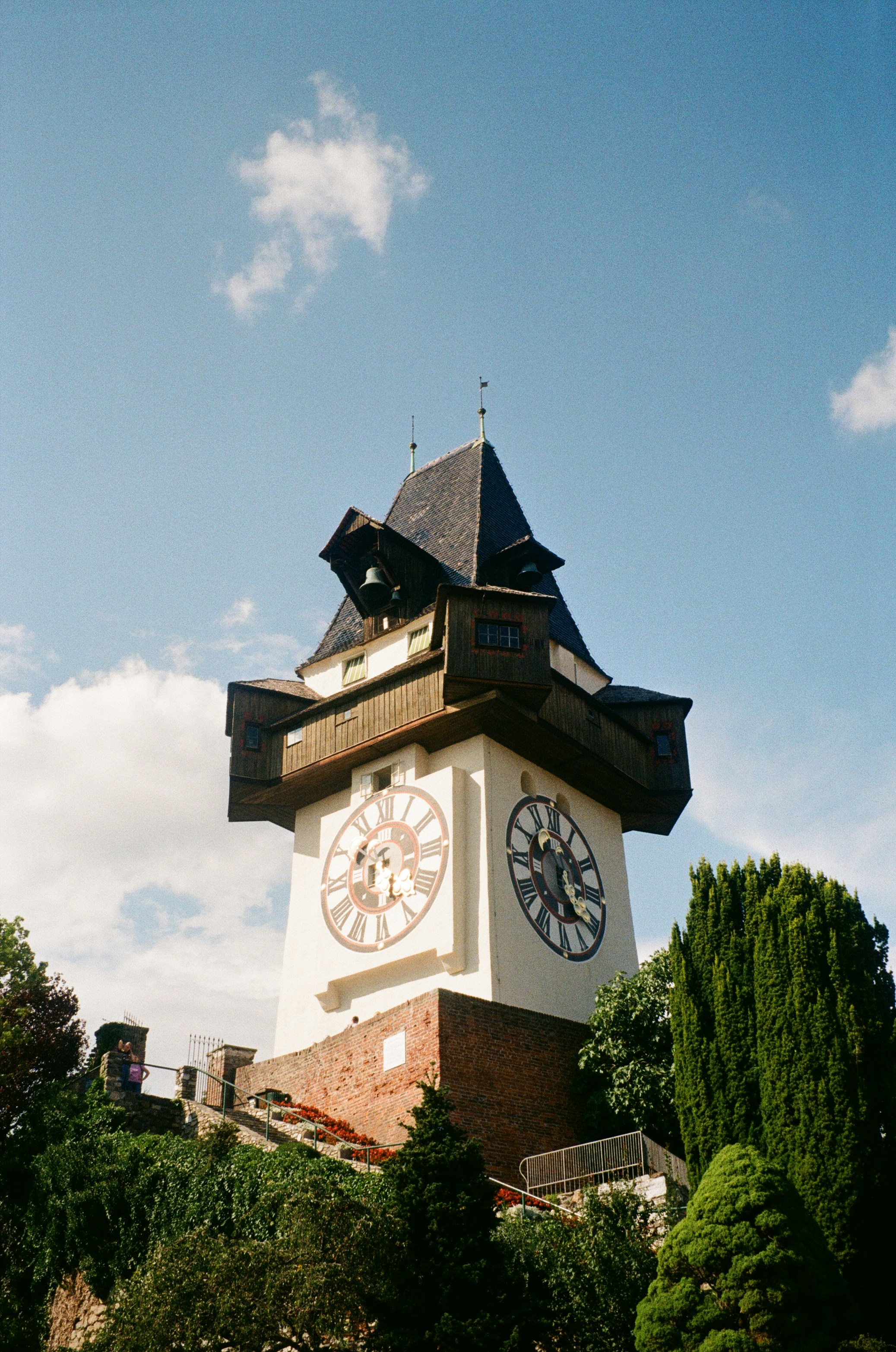 Historic clock tower with intricate clock faces and a wooden roof, surrounded by lush greenery and blue skies.