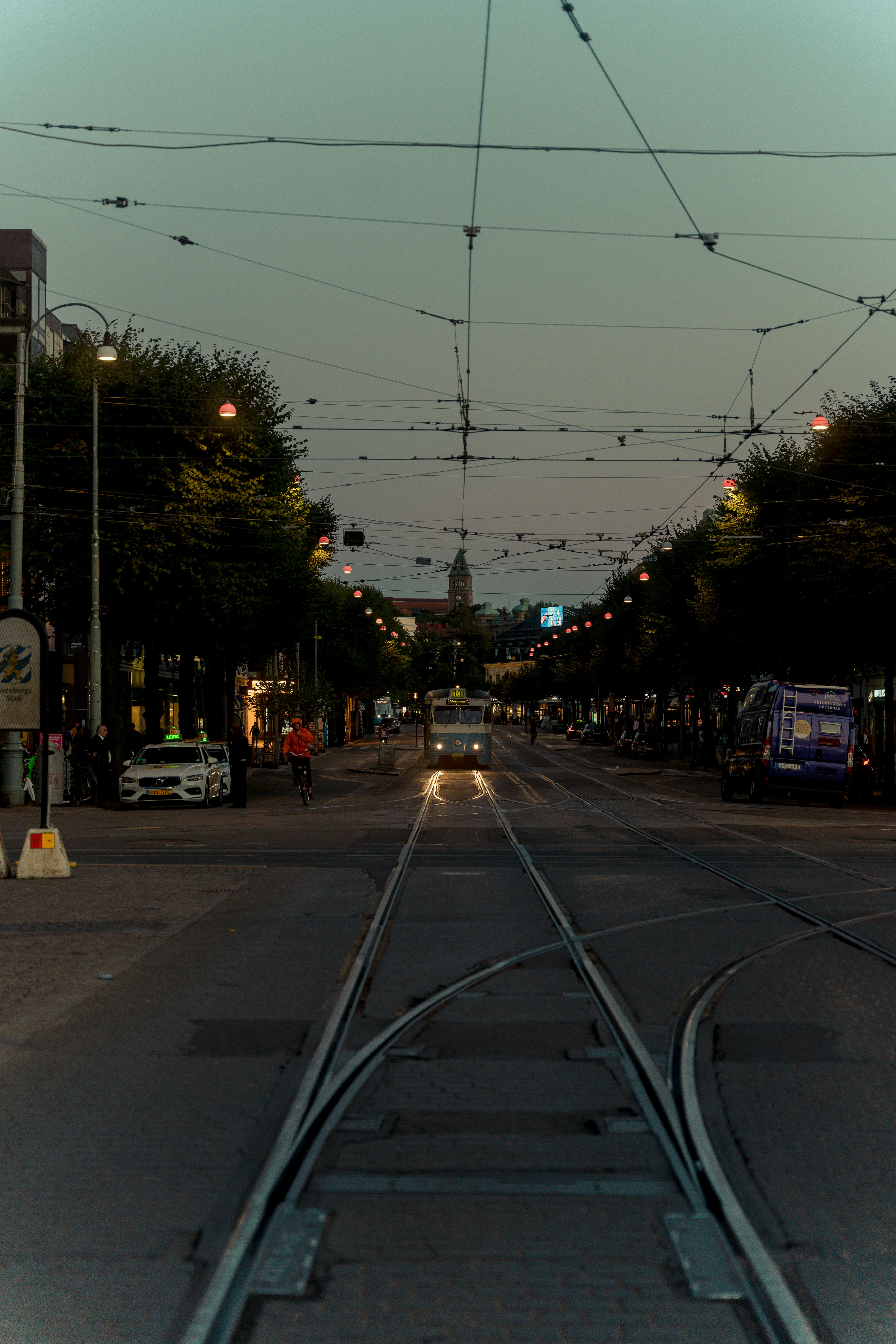 Tram gliding along tracks in a bustling urban street illuminated by soft evening lights, framed by trees and shops.