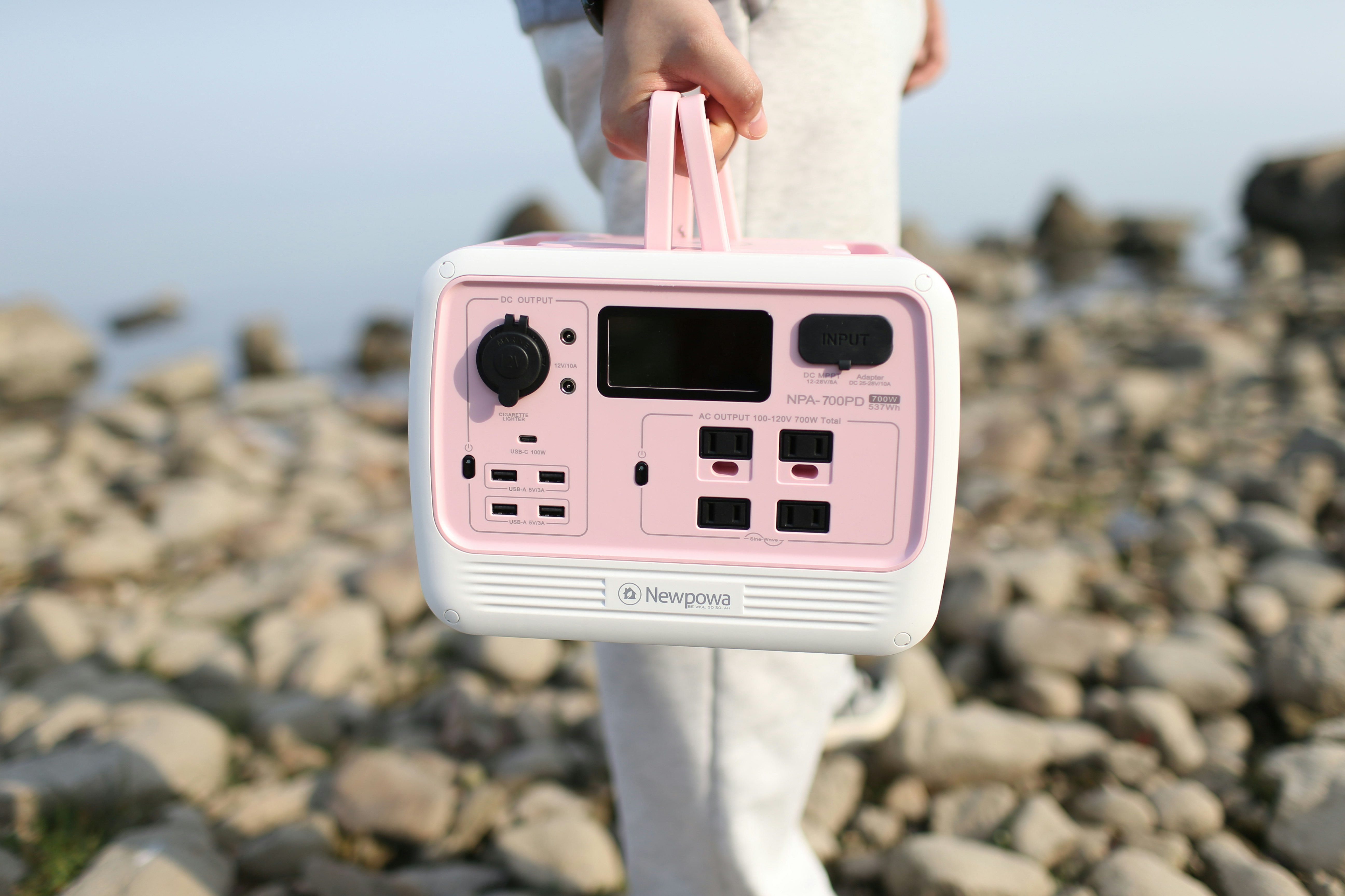 A person carries a pink Newpow power station while walking over rocky terrain by the water's edge.