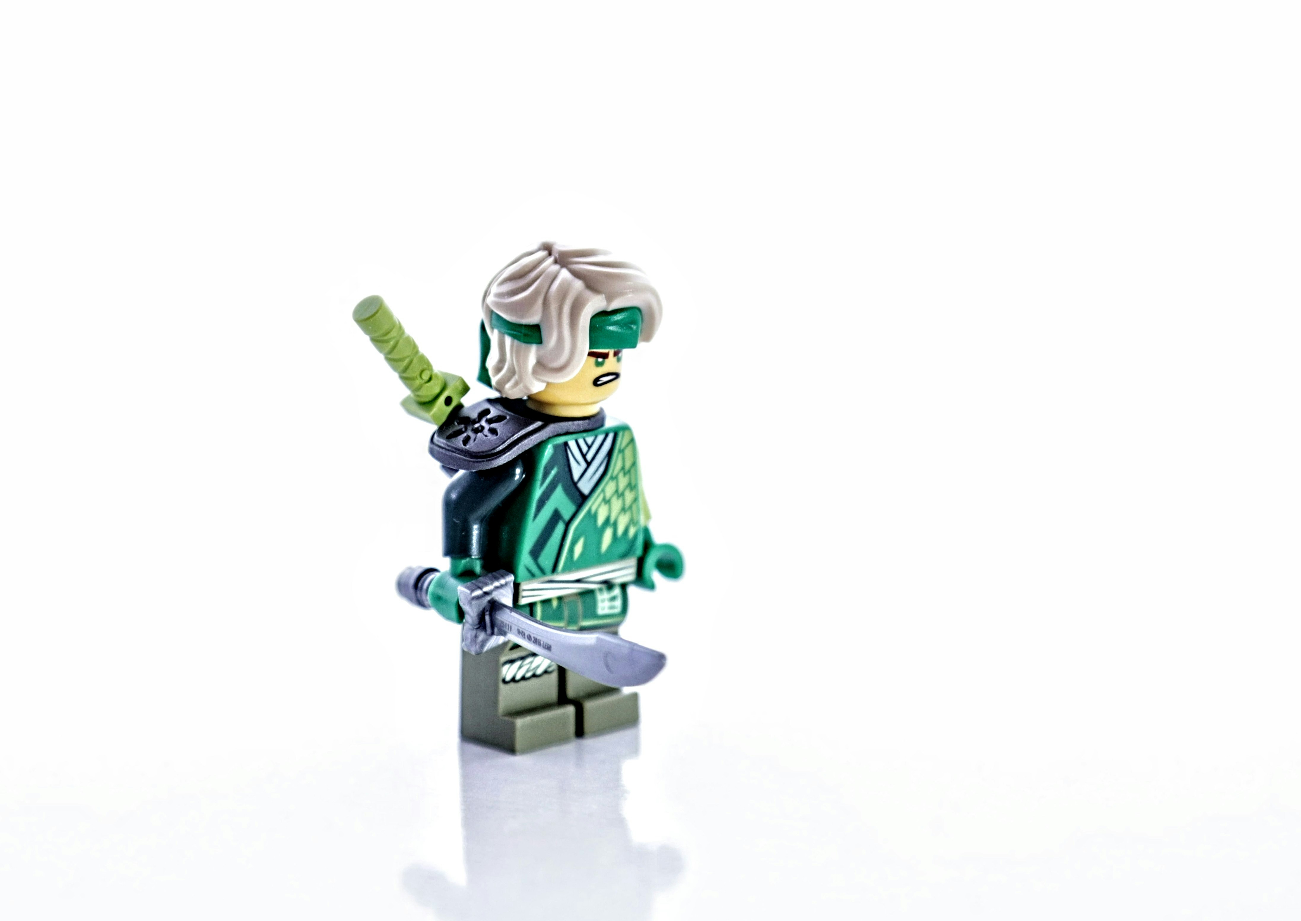 A single LEGO minifigure with green and teal attire stands on a glossy white surface, sword drawn and face partially covered by eyewear, isolated on a bright background.
