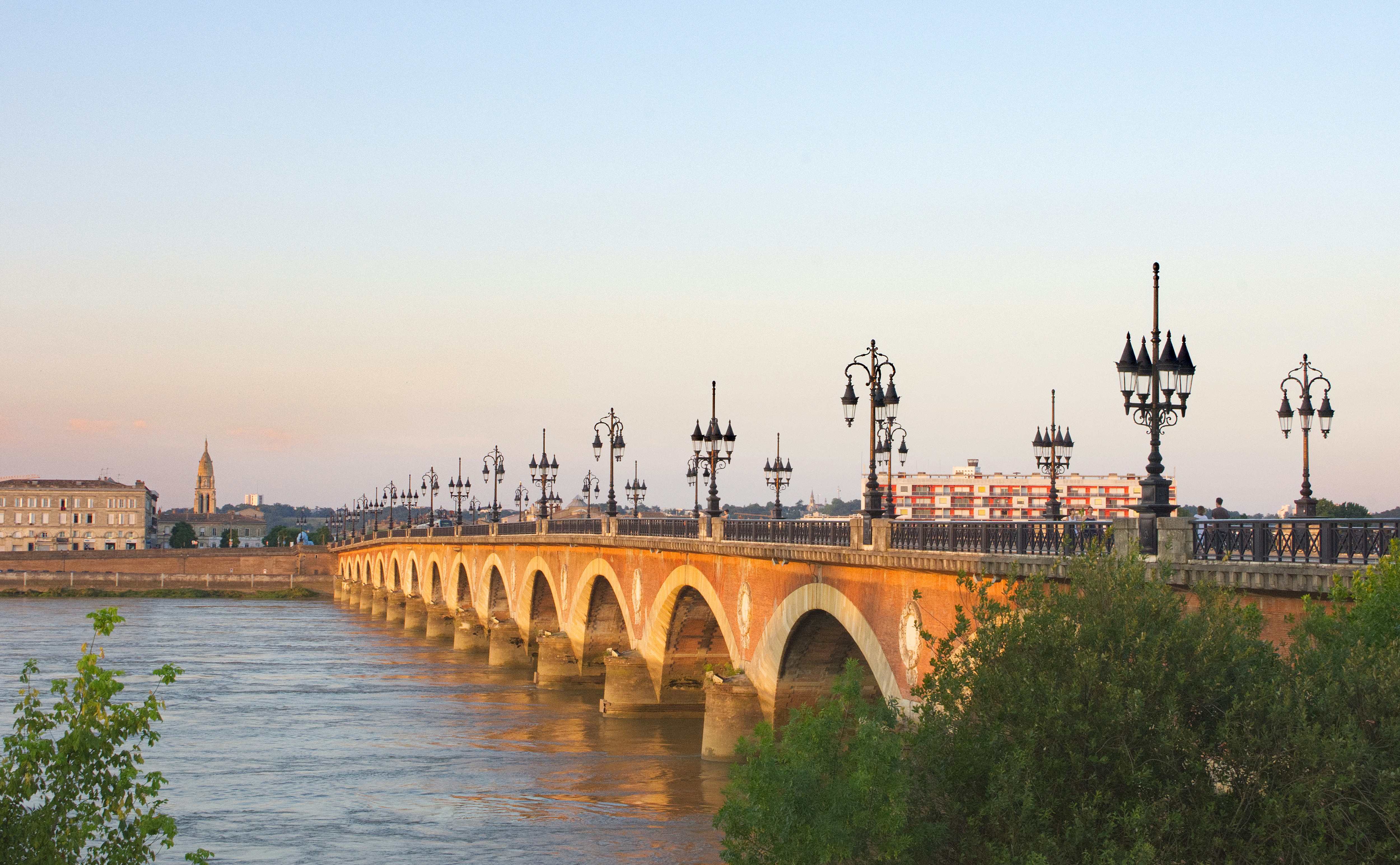 Historic bridge adorned with ornate street lamps spans a tranquil river, reflecting the soft hues of twilight. A serene urban landscape unfolds in the background.