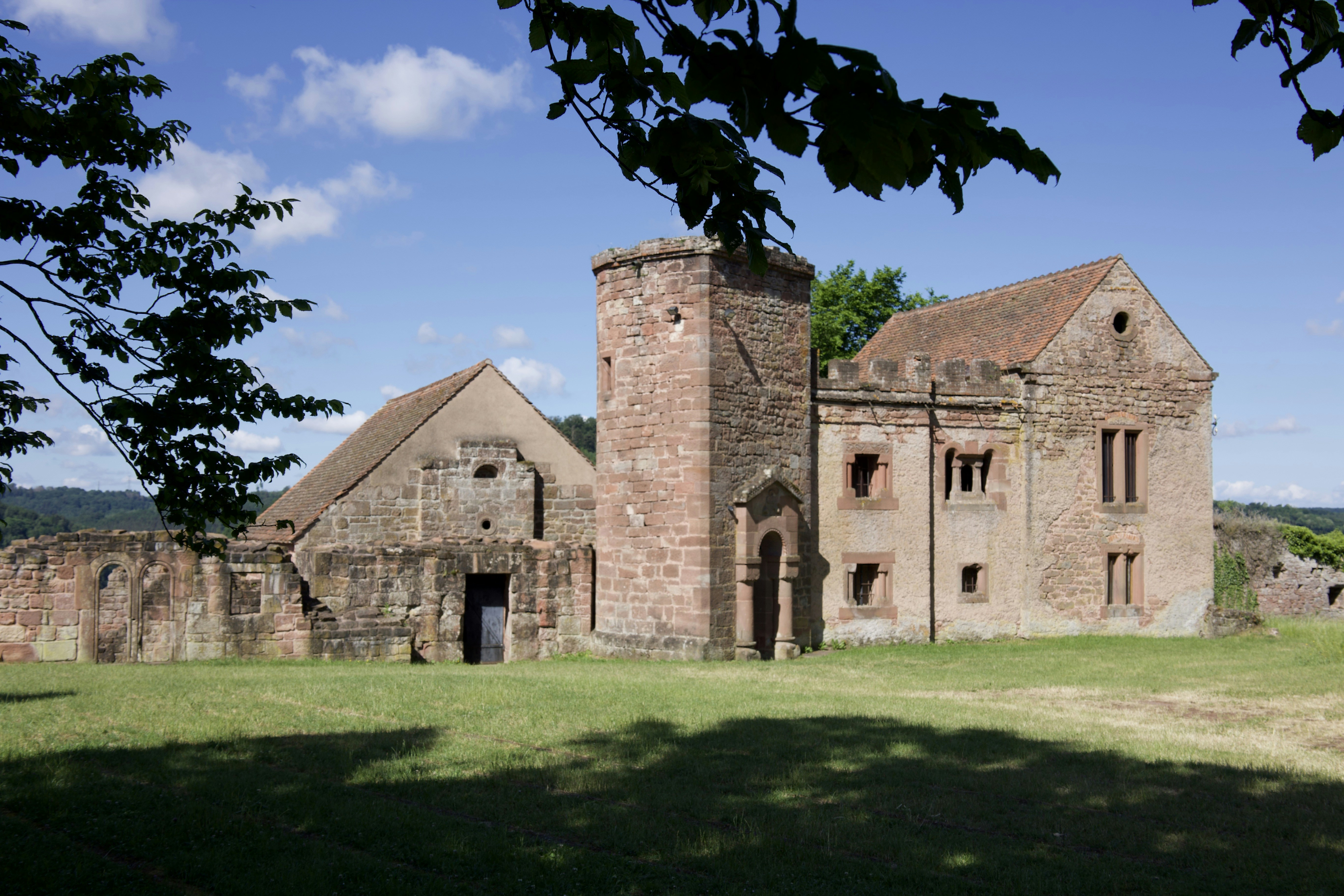 Medieval stone building with a prominent tower, surrounded by a grassy field and framed by tree branches.