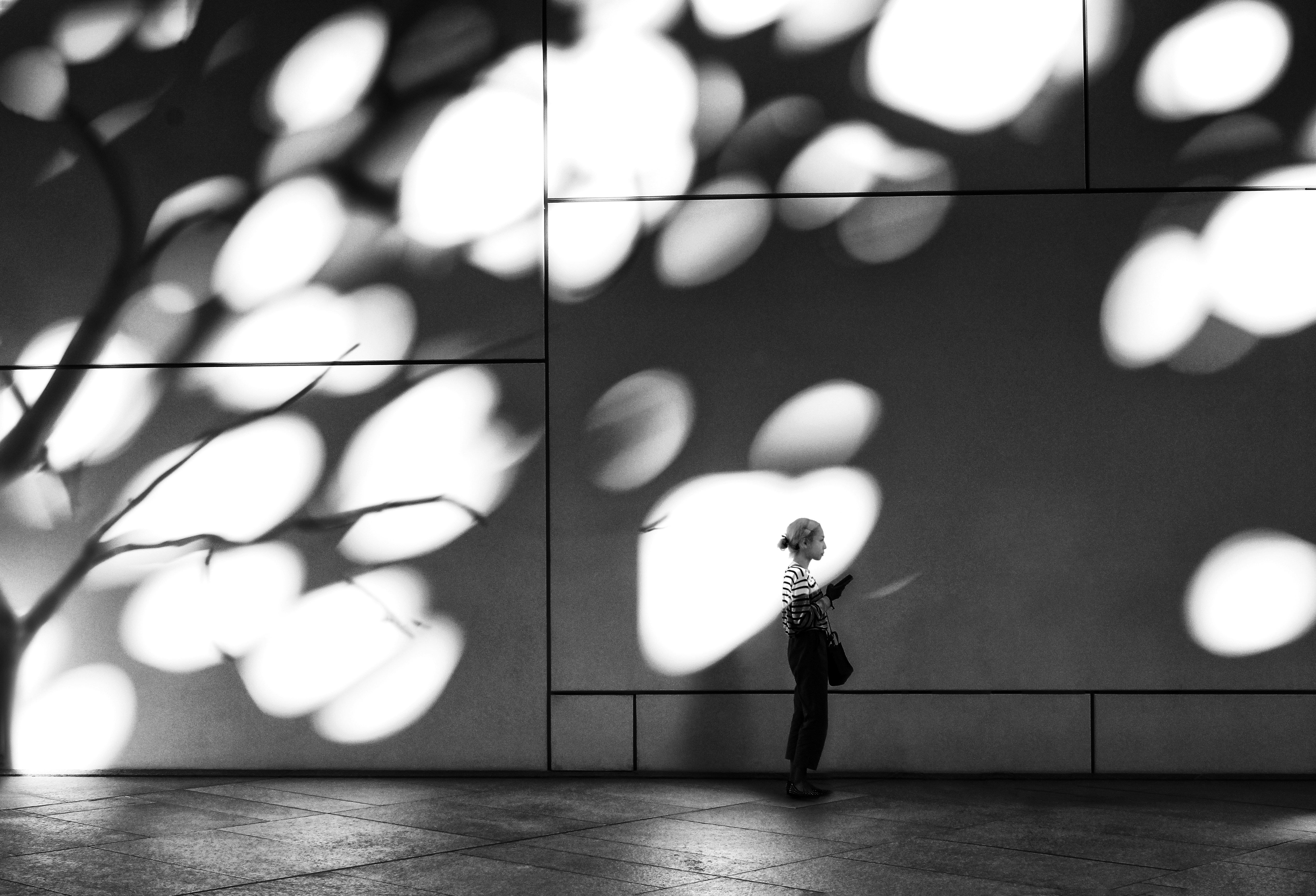 A solitary figure stands against a wall adorned with abstract shadows, creating a striking contrast between light and dark. The interplay of shapes evokes a sense of introspection.