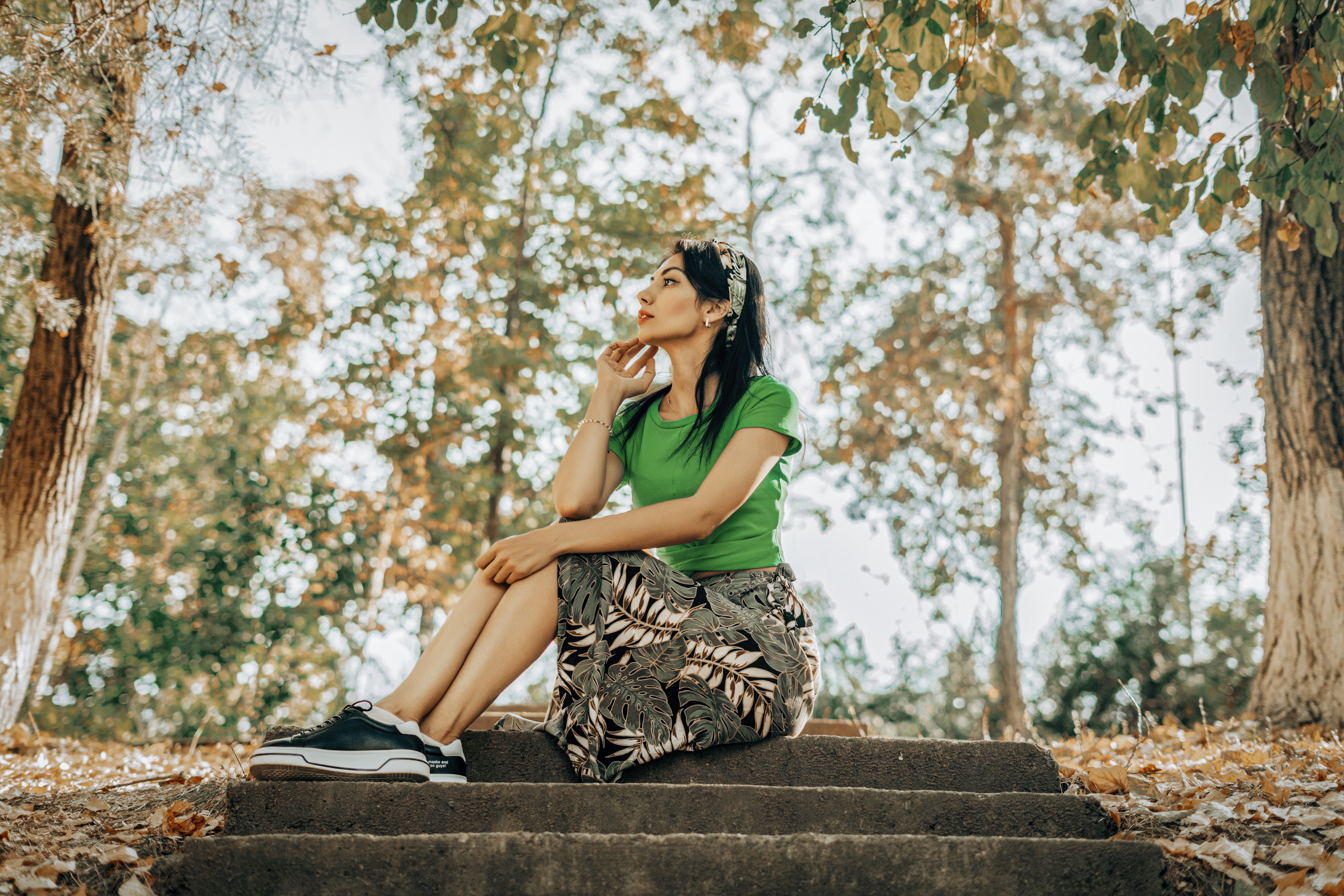 Woman in green top and patterned skirt sits thoughtfully on stone steps surrounded by autumn foliage.