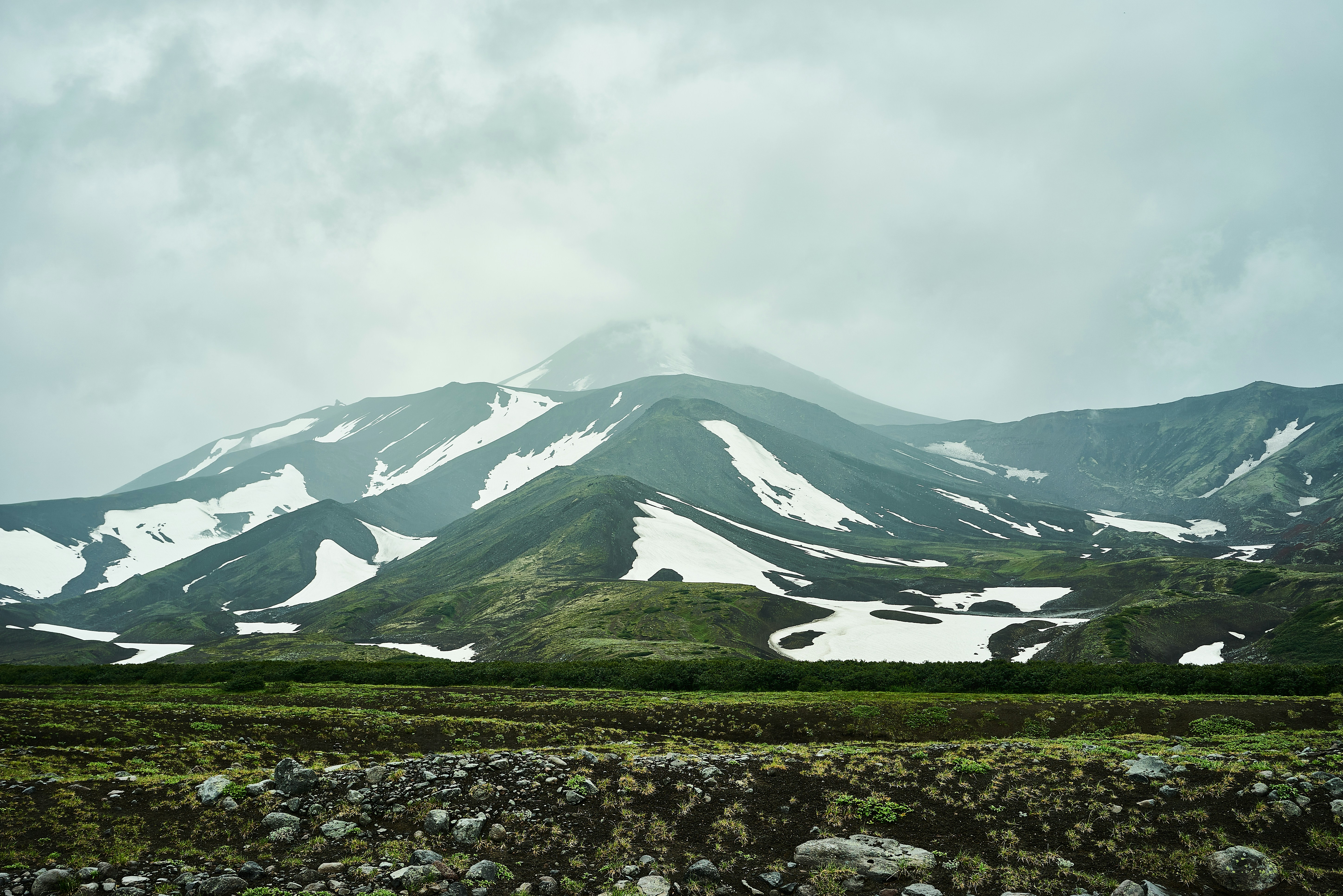 Lush green hills and snow-capped peaks under a cloudy sky showcase the raw beauty of a volcanic landscape. The scene captures the interplay of earth and sky.