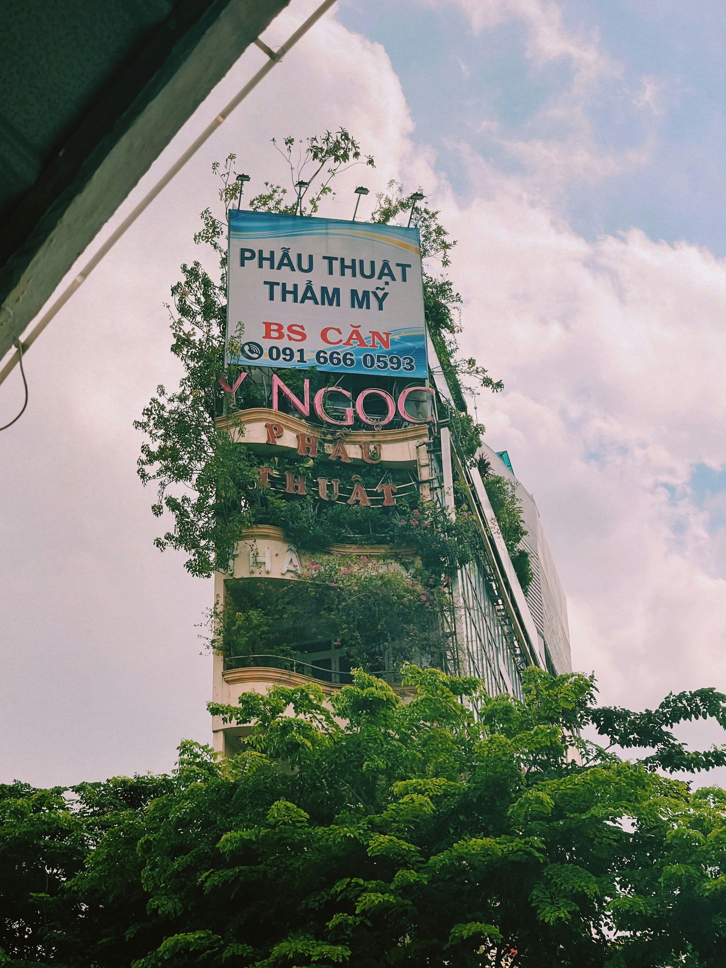 Tall building facade with a prominent sign reading 'PHẪU THUẬT THẨM MỸ' veiled by climbing greenery against a bright blue sky.