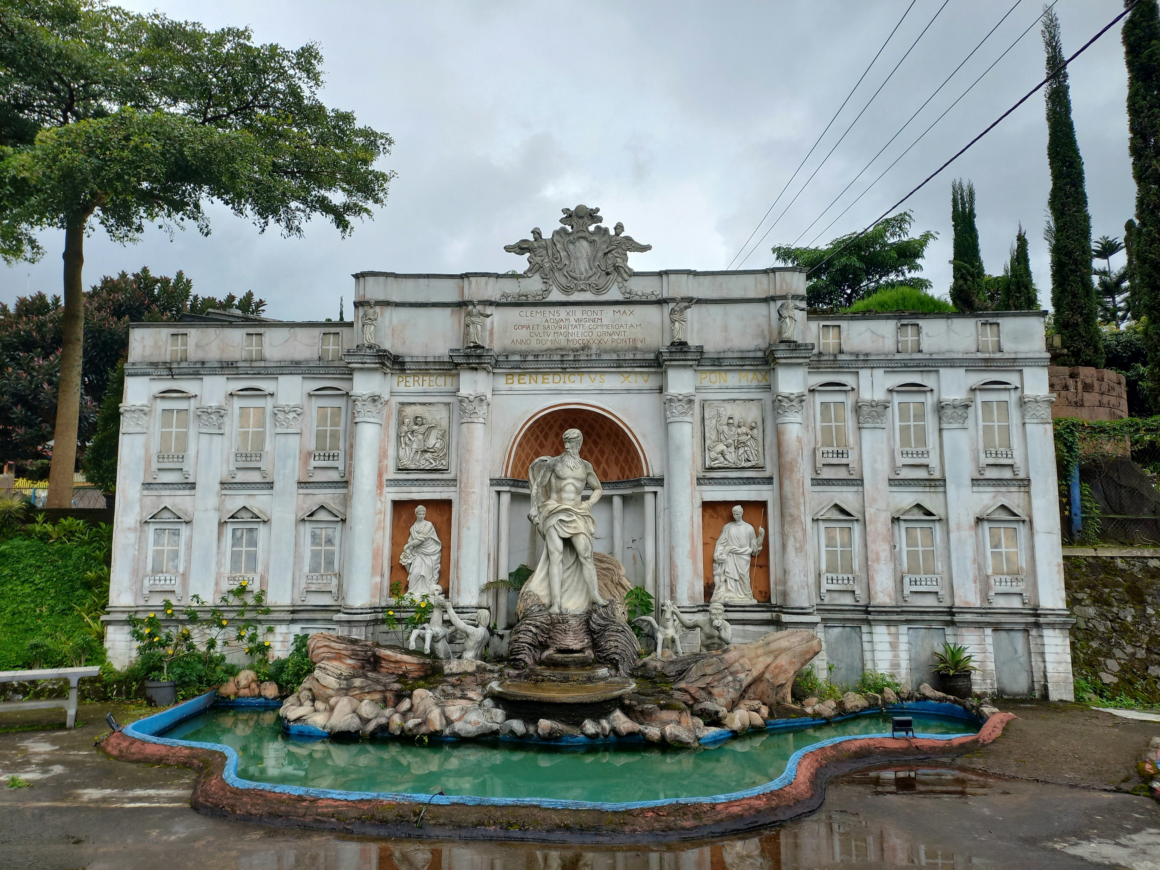 Ornate stone fountain in front of a classical architectural facade surrounded by greenery on a cloudy day.