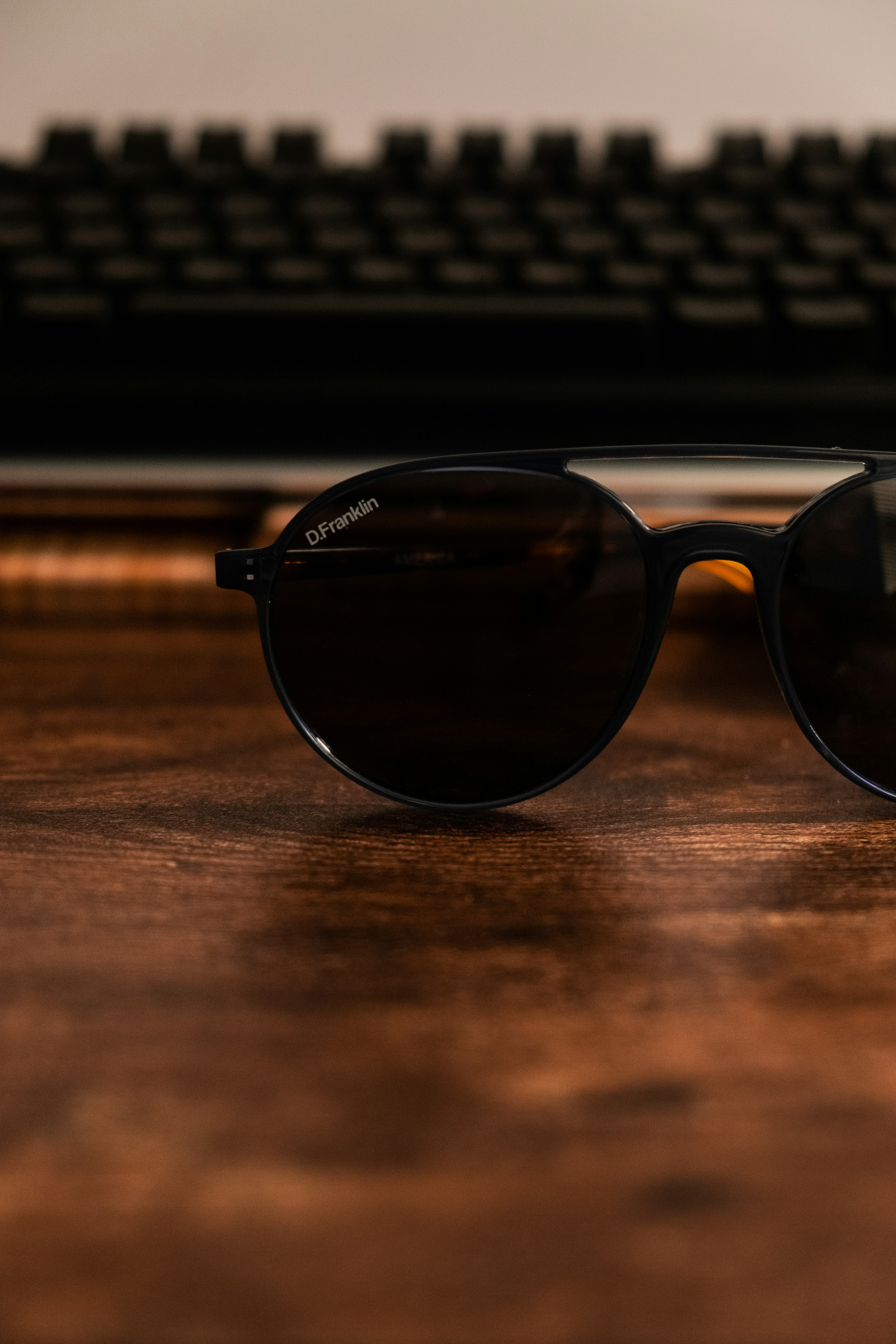 Round black sunglasses resting on a wooden surface with a keyboard blurred in the background.