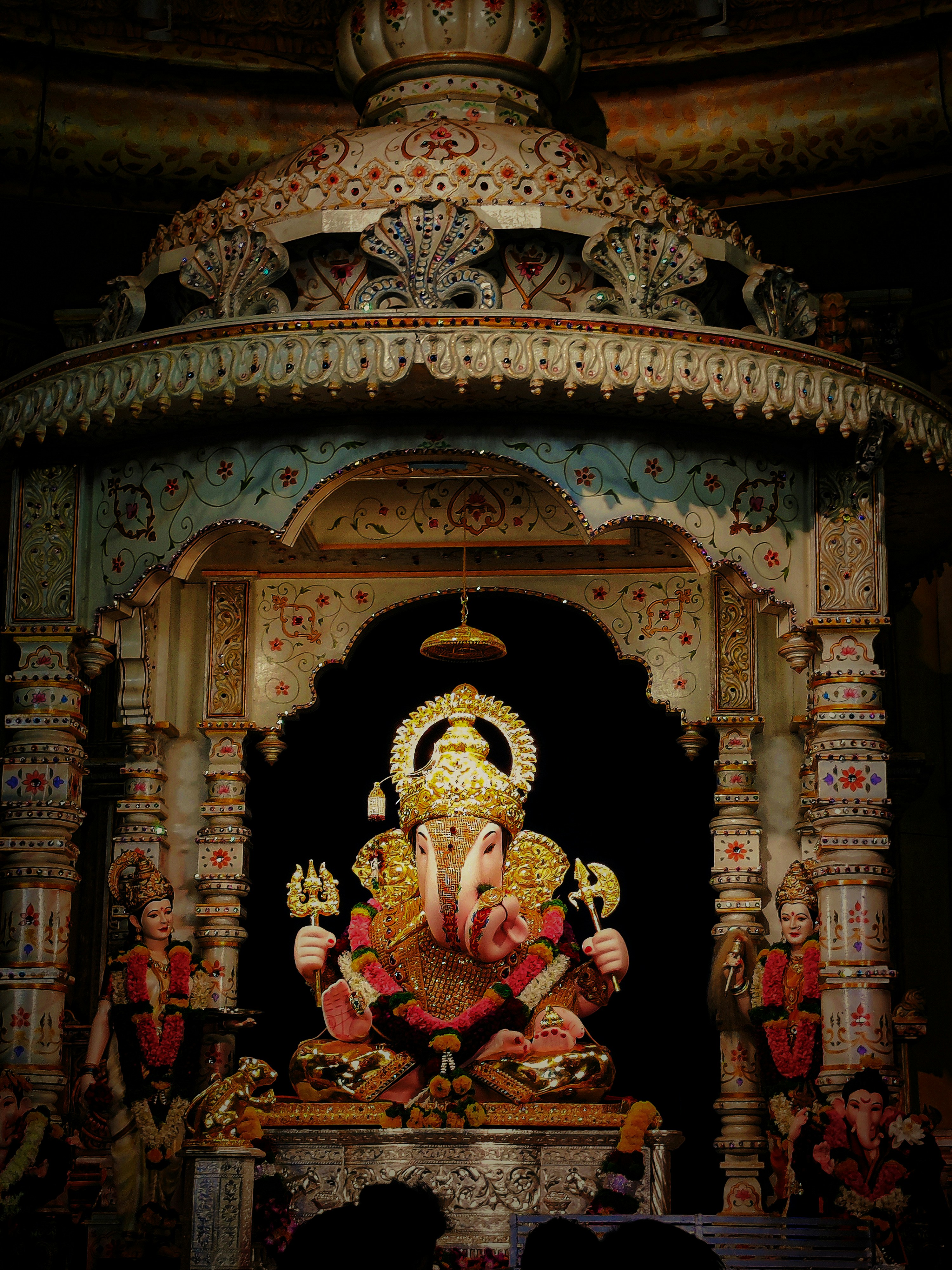 Ornately decorated idol of Lord Ganesh seated under a richly adorned canopy, surrounded by floral offerings and devotees in a dimly lit setting.