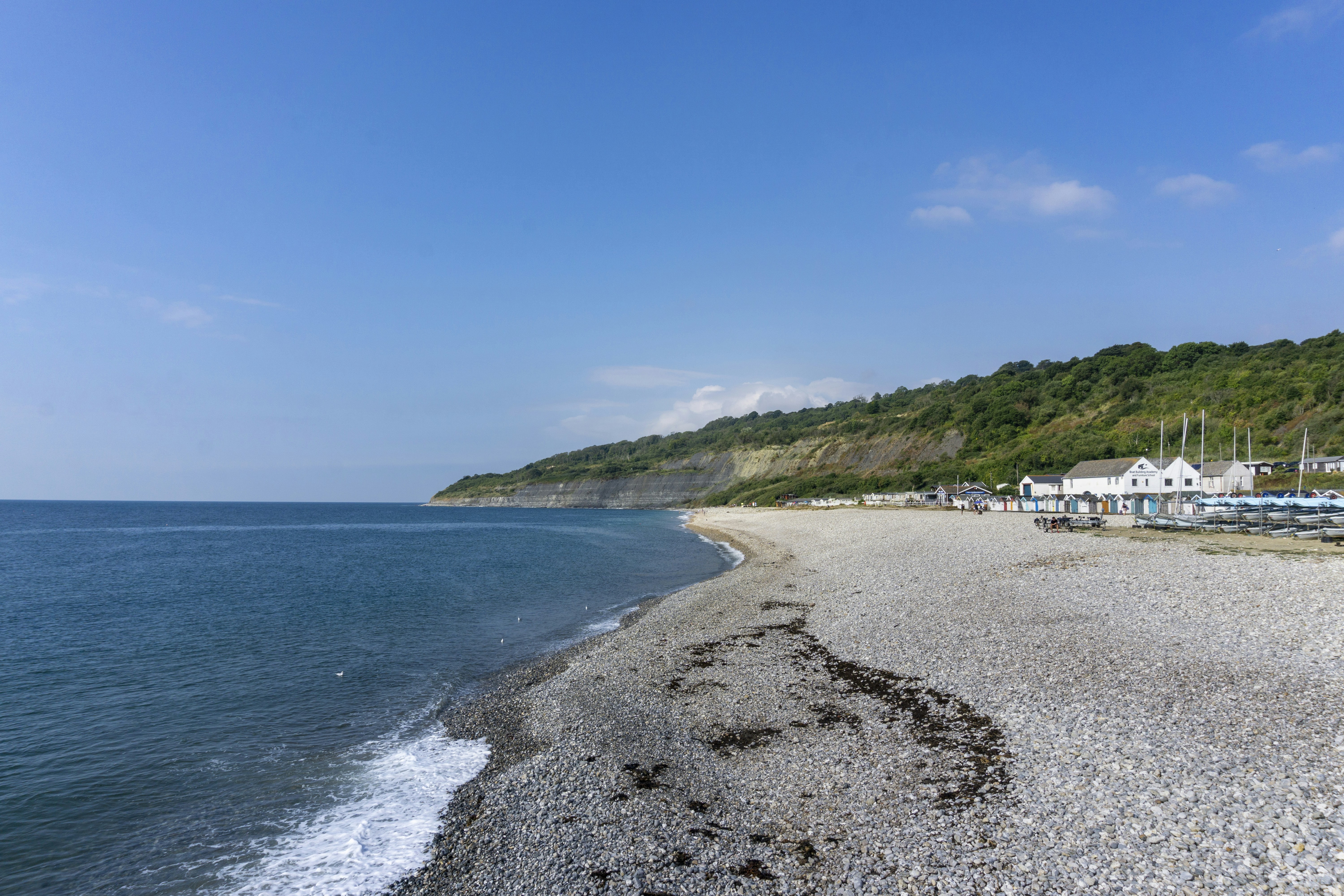 Pebble beach meeting calm blue waters under a clear sky, with a distant hillside and small buildings along the shoreline.