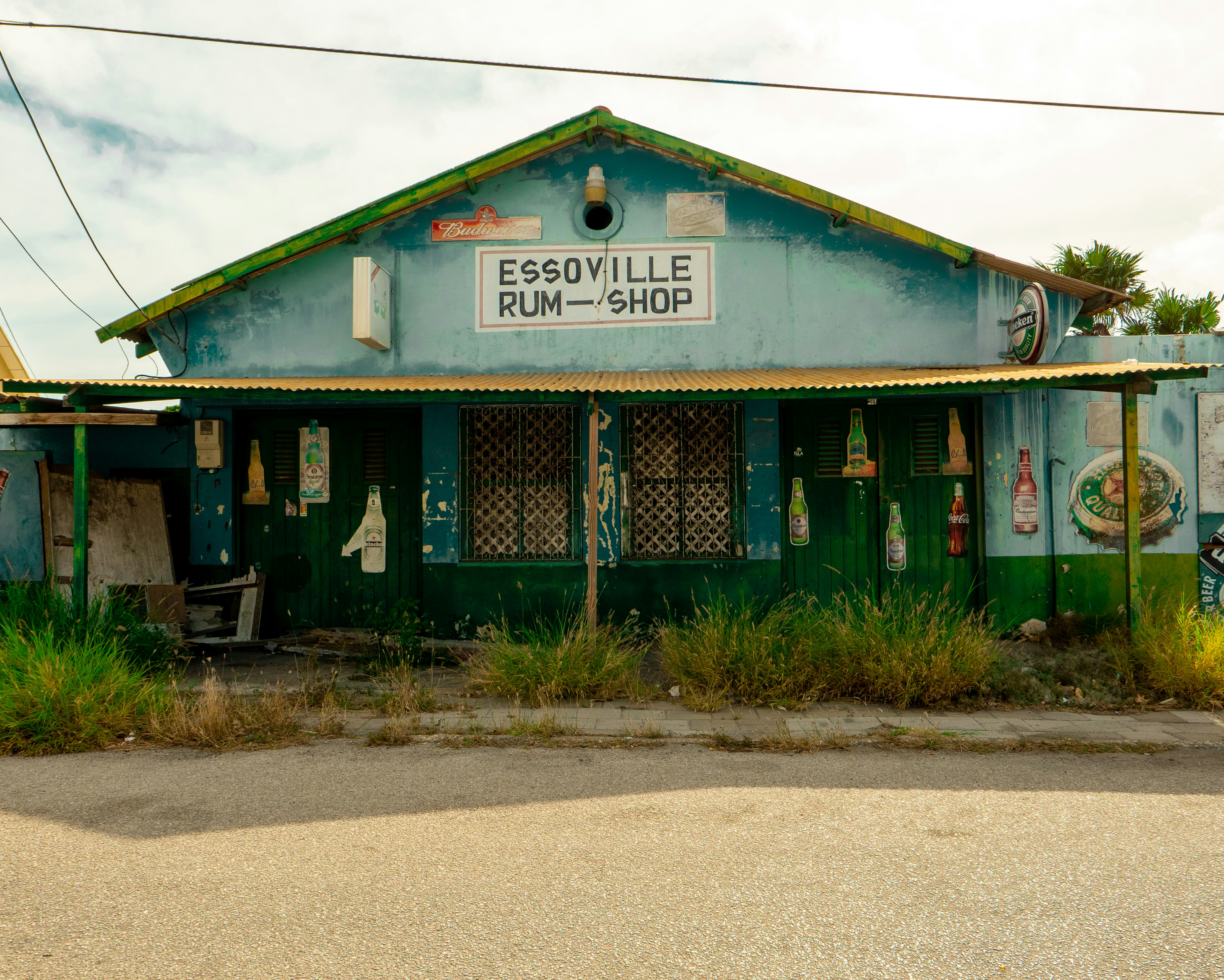 Weathered blue storefront with a corrugated roof and the sign 'Essoville Rum Shop' along a quiet road, flanked by overgrown grass and wall posters.