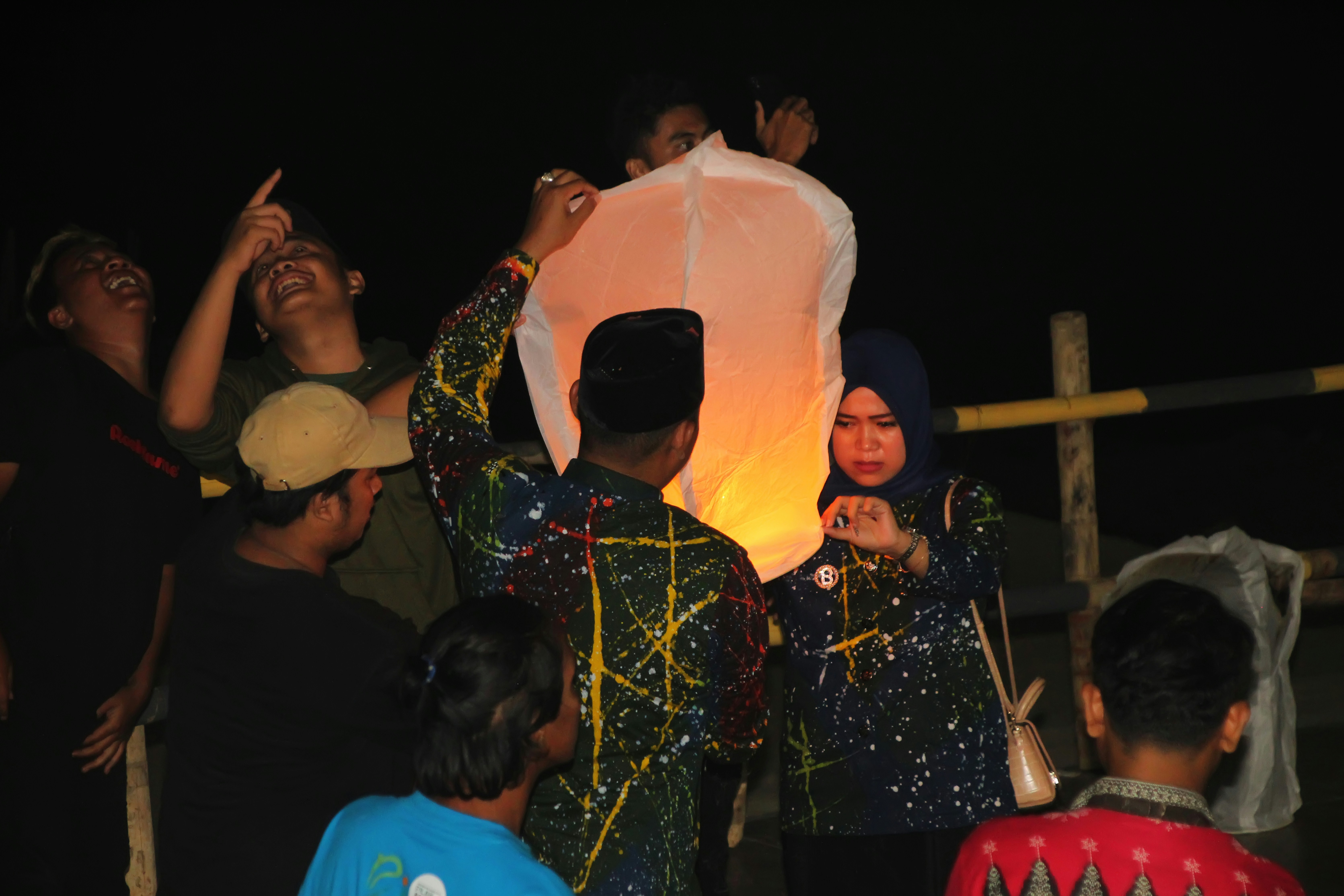 Group gathers to release a glowing lantern into the night sky, surrounded by darkness.