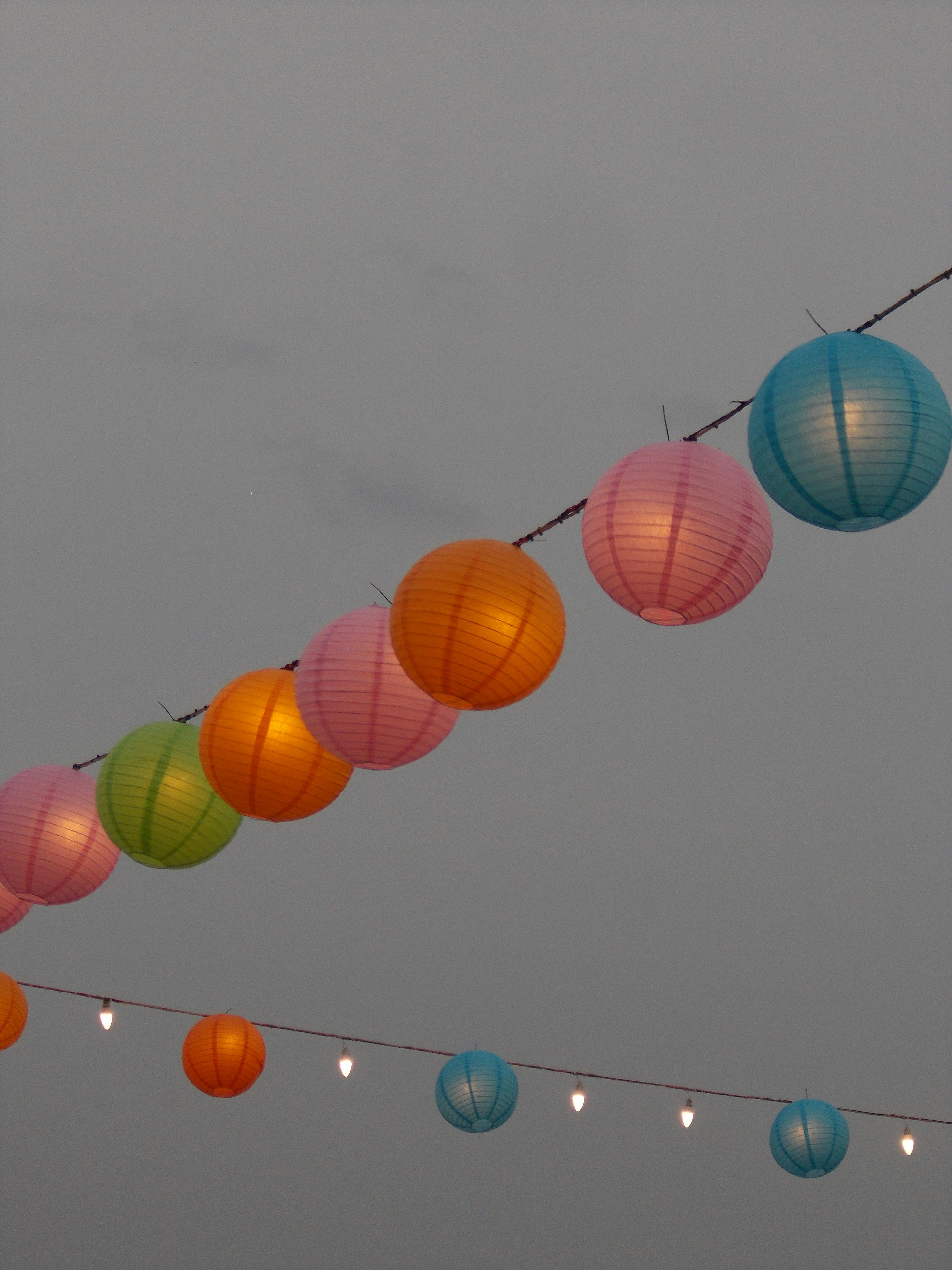 Colorful paper lanterns on a wire stretch diagonally across a muted gray sky. A straightforward string-light moment captured as a photograph.