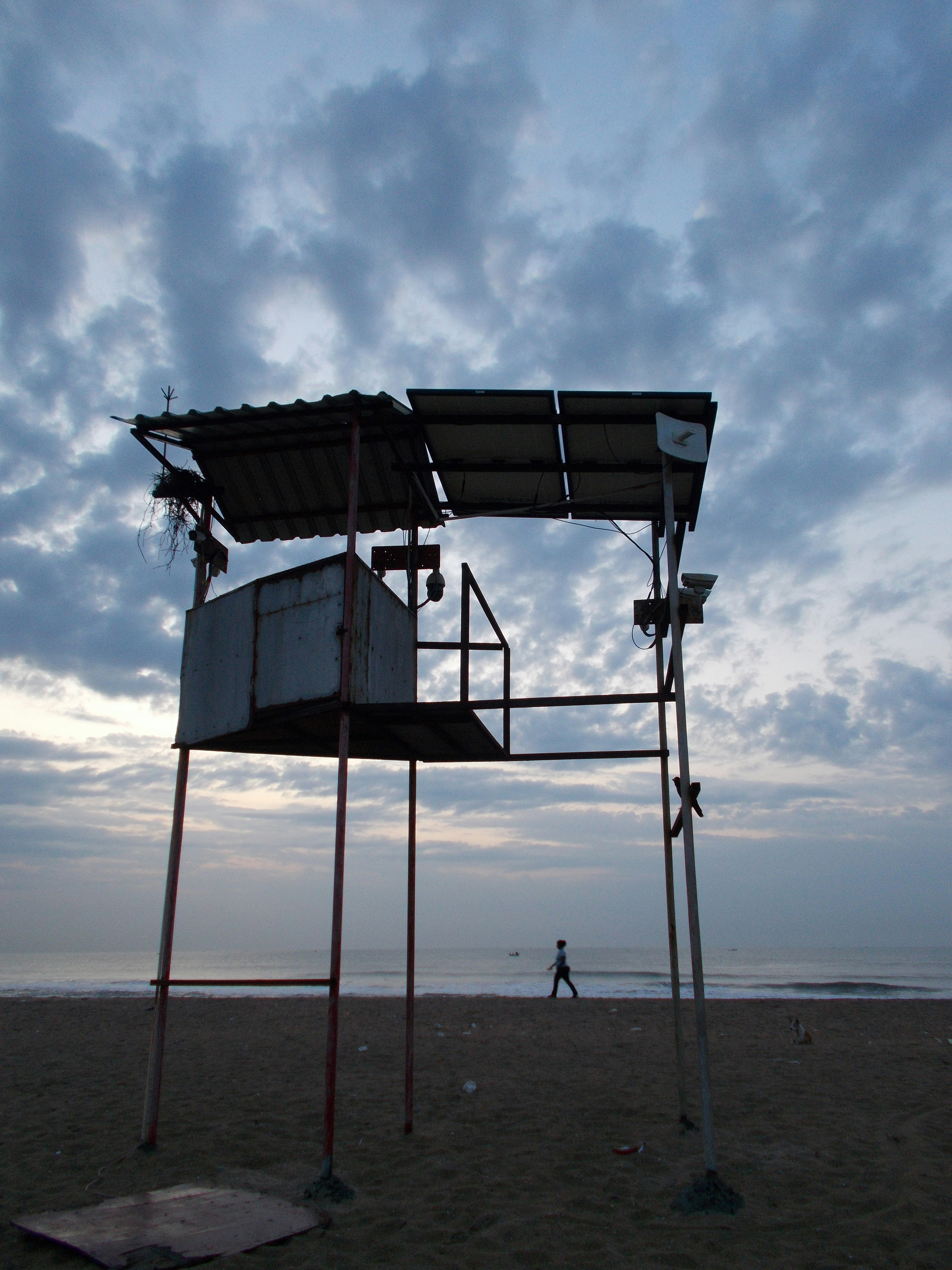 A weathered lifeguard tower stands silhouetted against a cloudy sky at twilight, while a solitary figure strolls along the beach. 