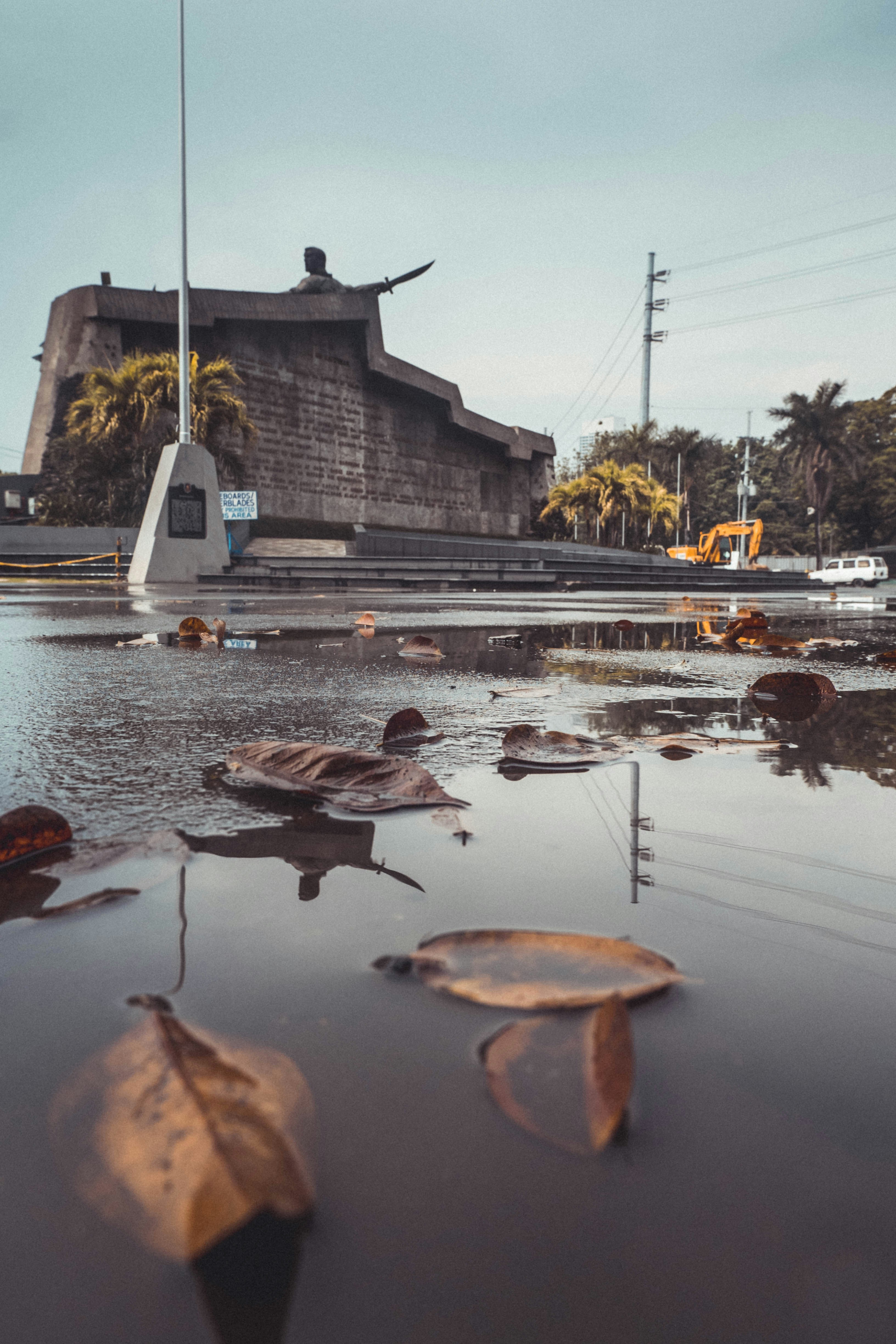 A large stone monument topped with a winged sculpture dominates a flooded urban plaza; water pools around its base. Reflections shimmer on the surface as leaves drift in the foreground, capturing a cityscape suspended in rain.