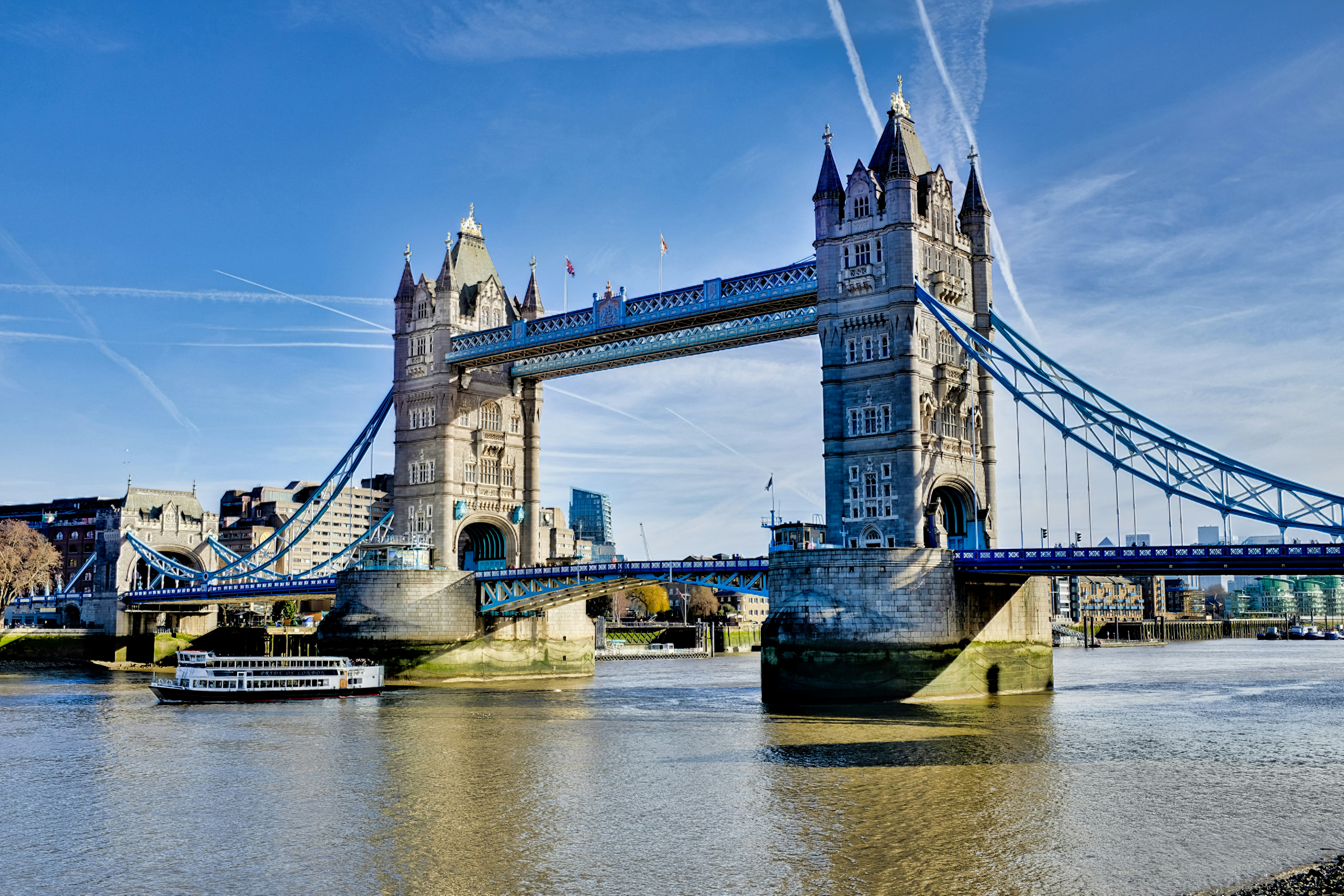 Tower Bridge stands proudly over the River Thames, showcasing its iconic architecture against a clear blue sky. A boat glides beneath the bridge, adding a touch of life to the scene.