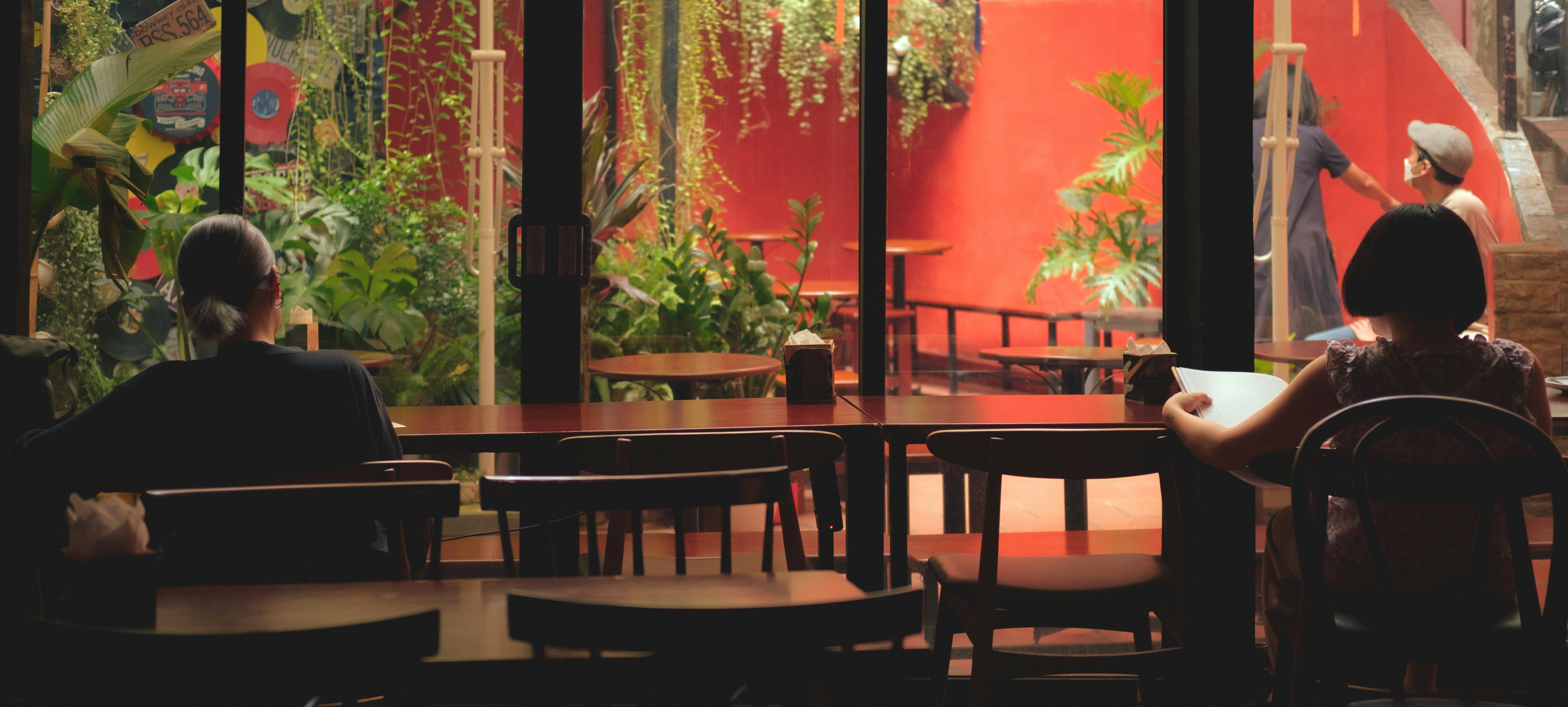 Two patrons seated in a cozy café, surrounded by vibrant greenery and a striking red wall. The scene captures a serene atmosphere with a hint of urban life.