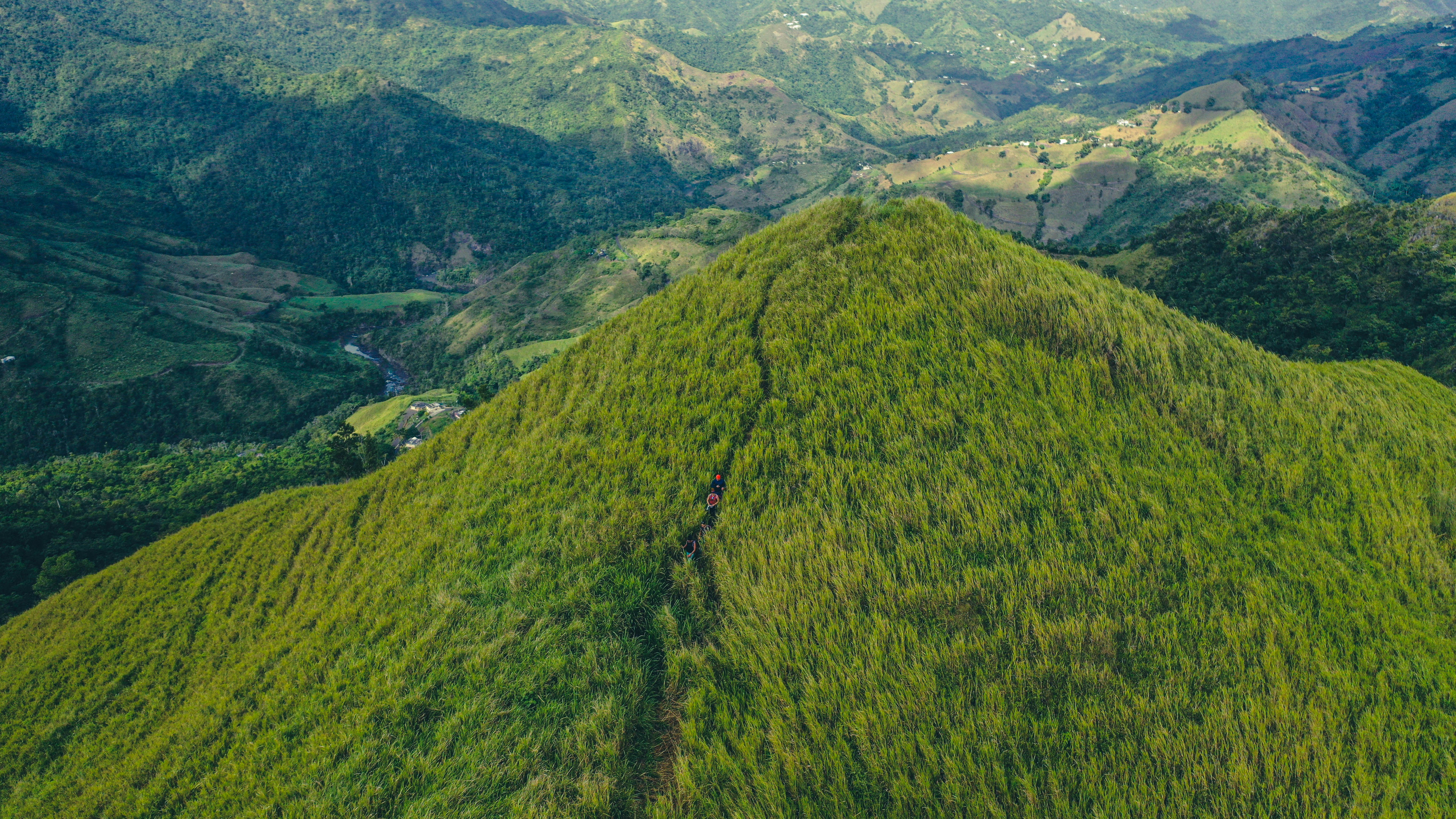 Aerial view of a lush green mountain peak with a winding trail leading up, surrounded by rolling hills and valleys.